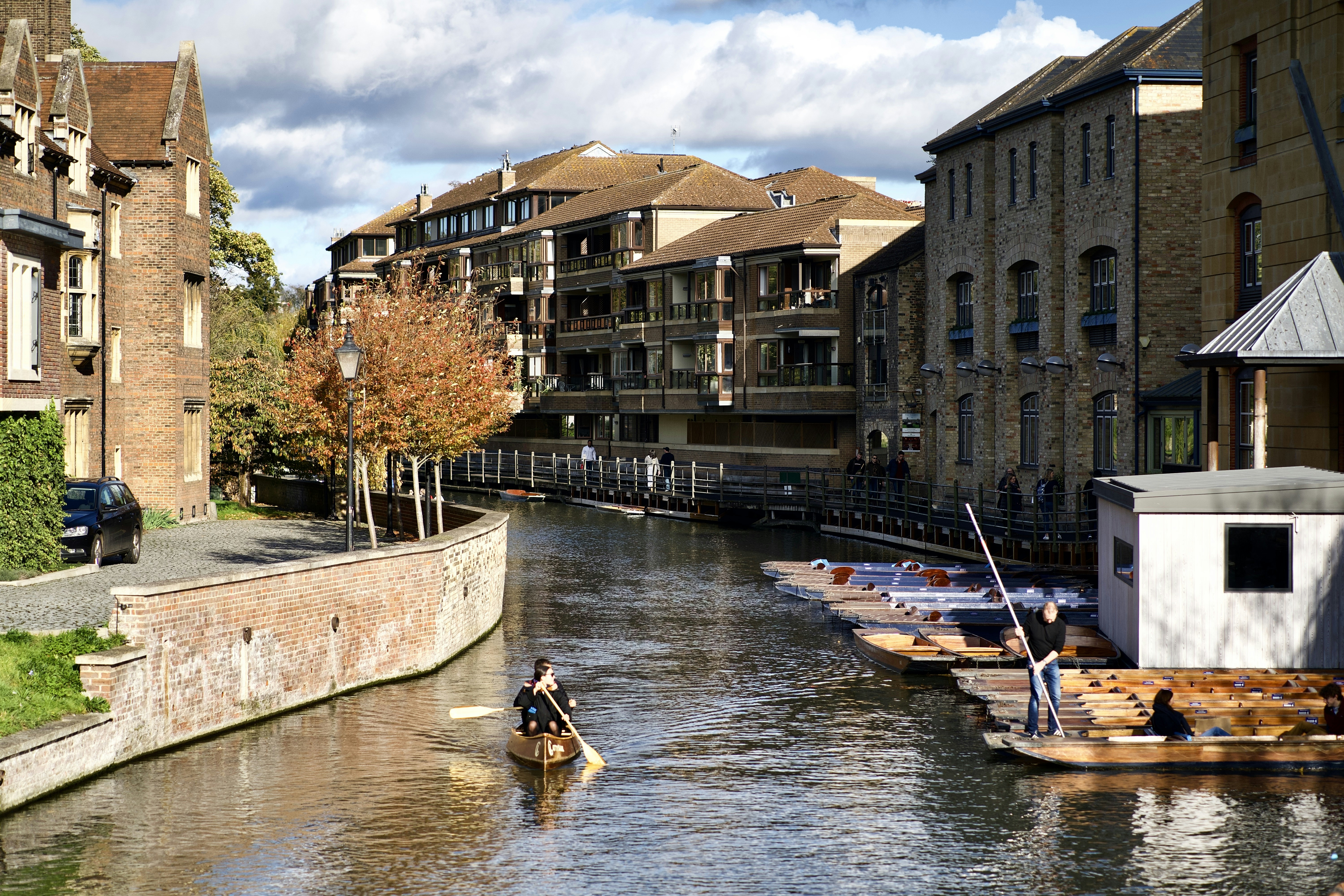 A lone rower navigates a narrow canal flanked by historic brick buildings under a partly cloudy sky.