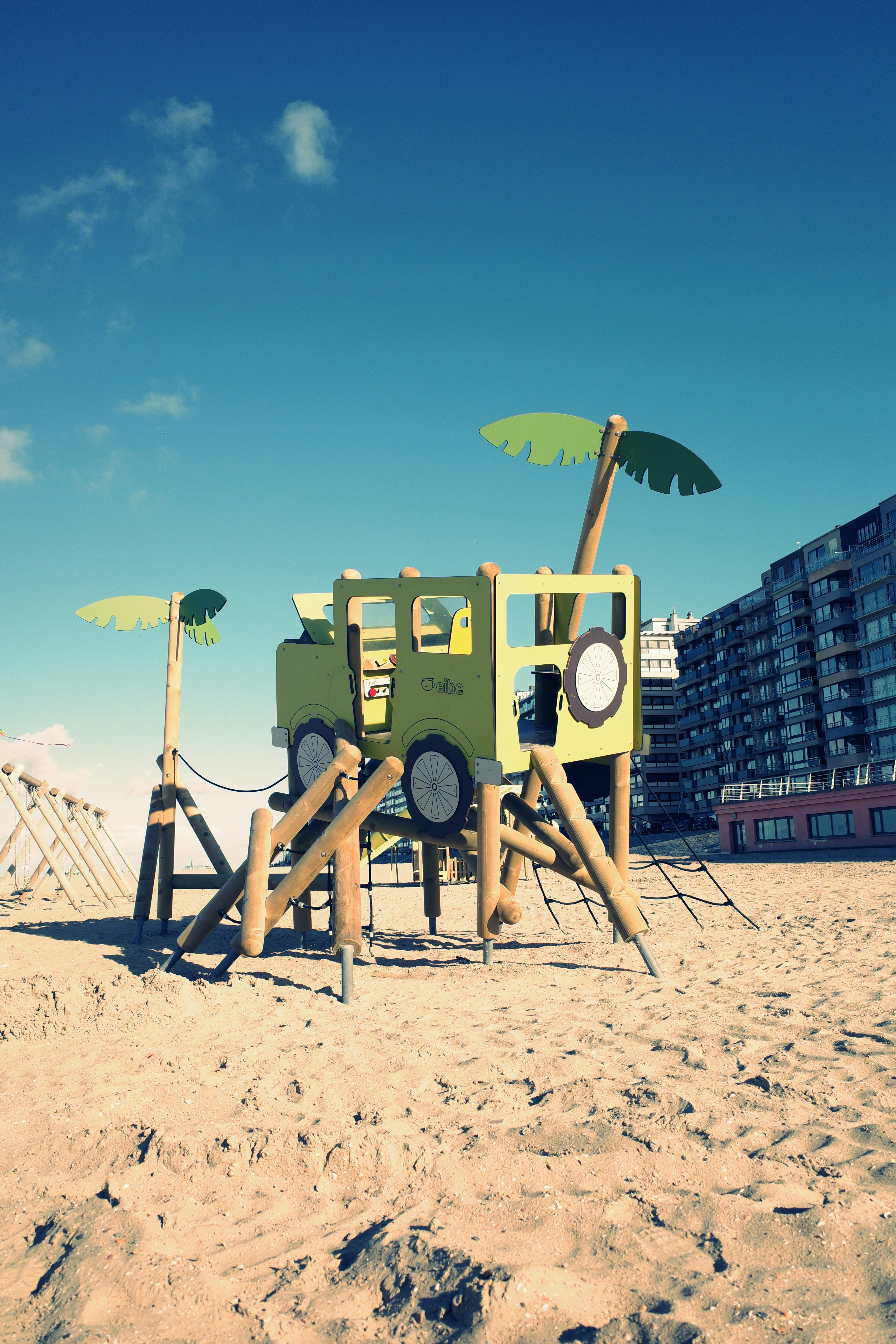 Shoreline Jungle Gym: Yellow-Green Playground on Sunlit Sand with an Urban Backdrop