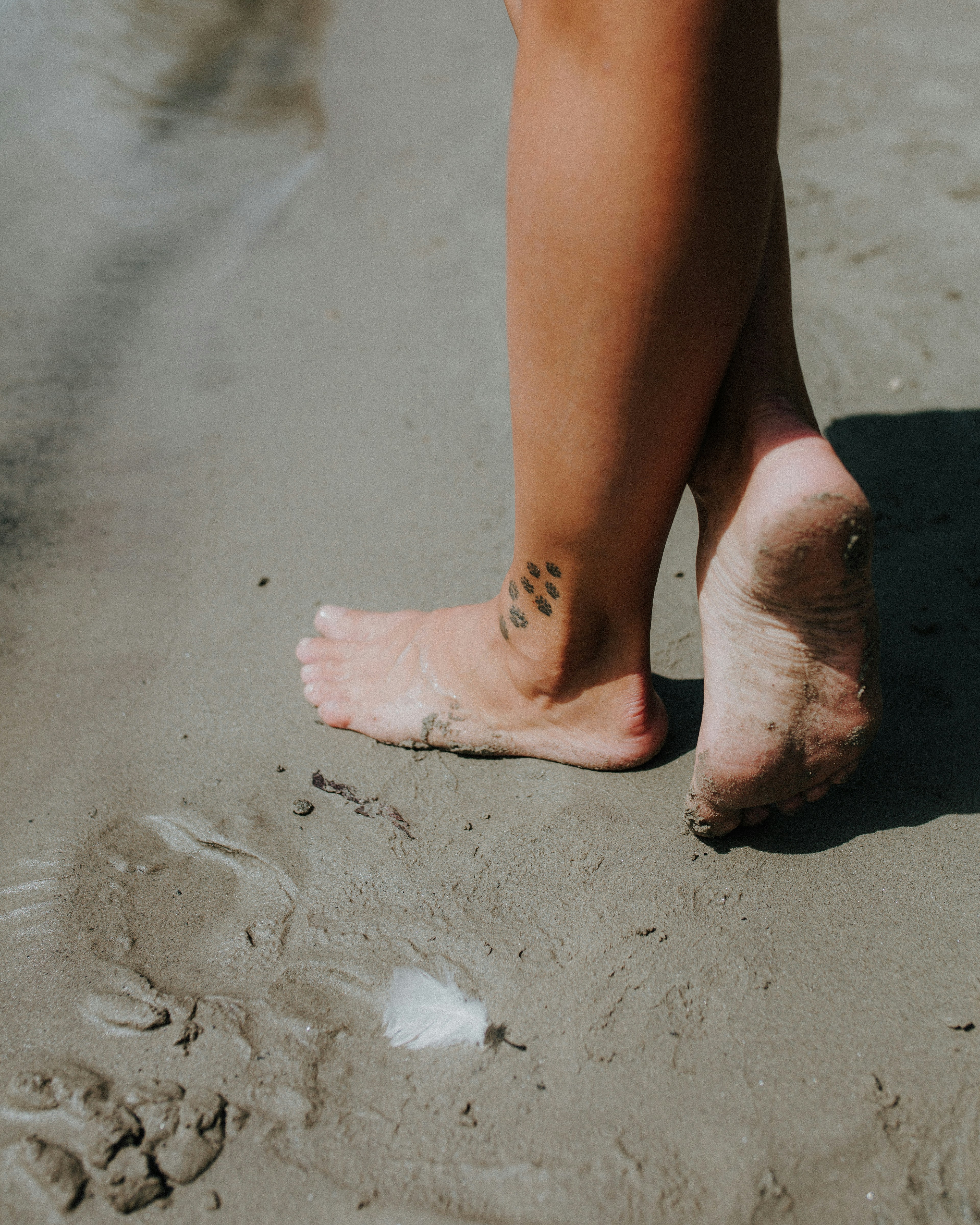 A person's feet, adorned with tattoos, tread softly on a sandy beach, with delicate traces of water nearby. The scene captures a moment of connection with nature.