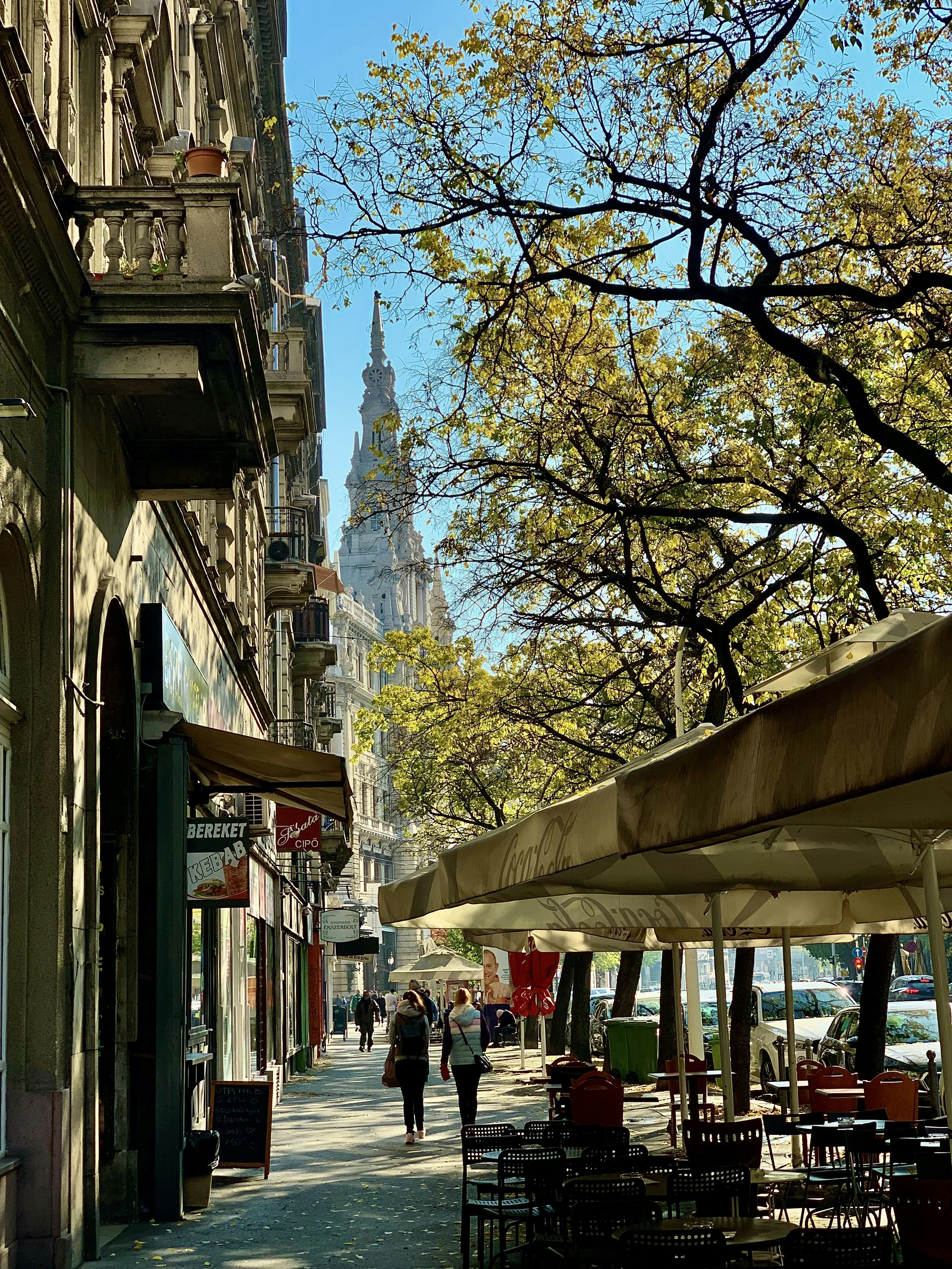 Charming street scene featuring outdoor dining and historic architecture, with a tall spire peeking through lush trees. 