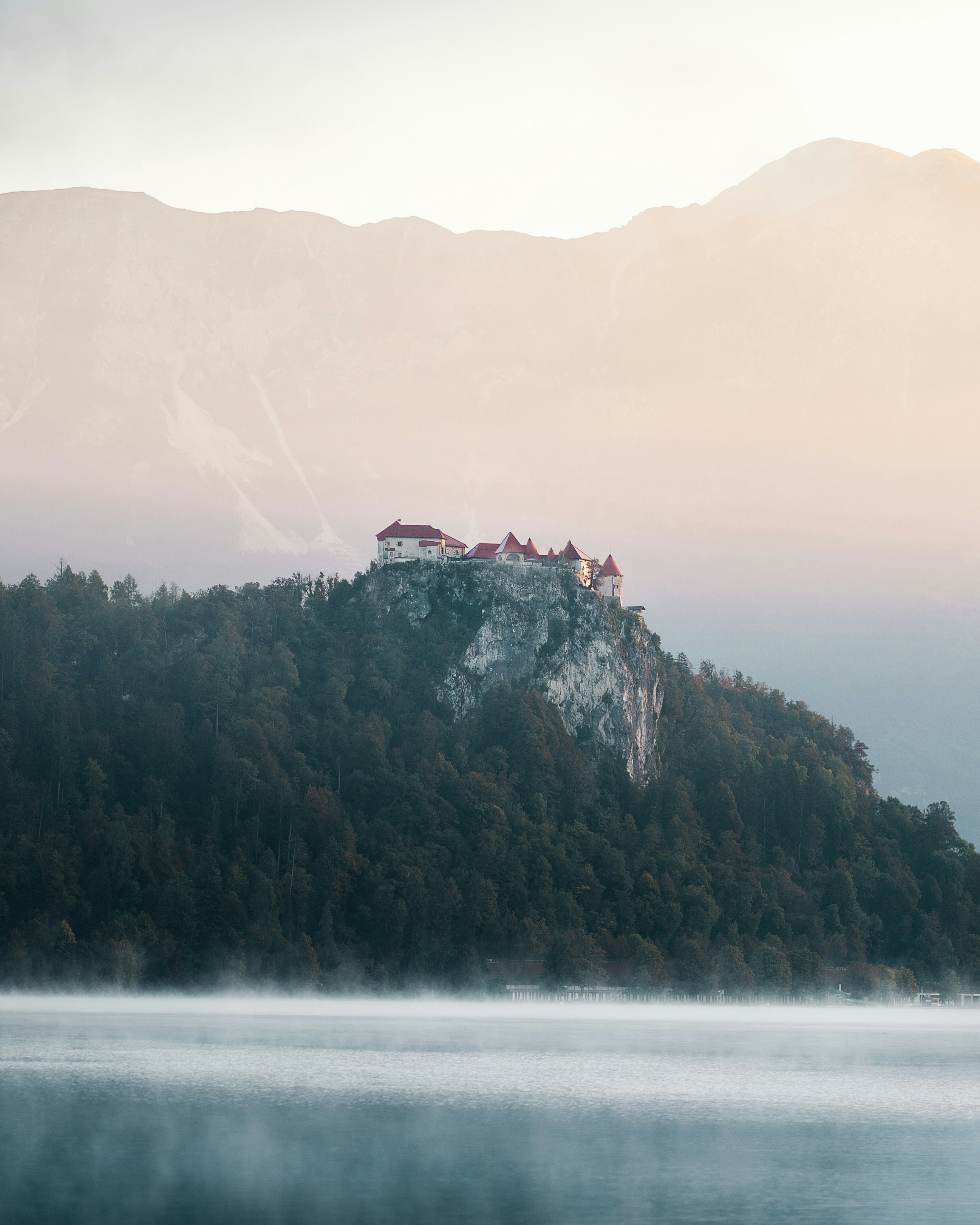 Castle perched atop a forested cliff with mist rising from the tranquil lake below.