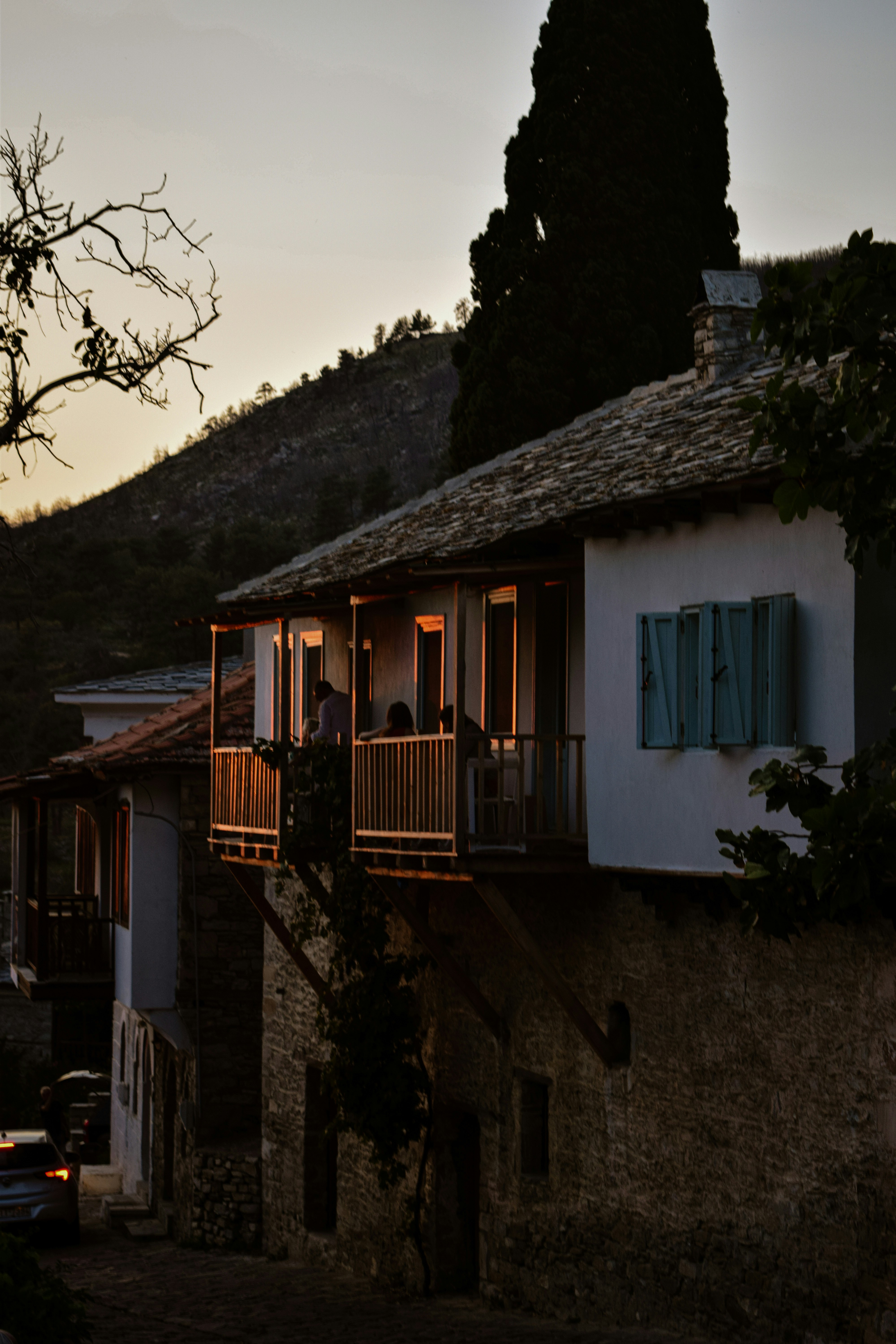 Charming village houses with illuminated balconies at dusk, framed by a mountainous backdrop and silhouetted trees.