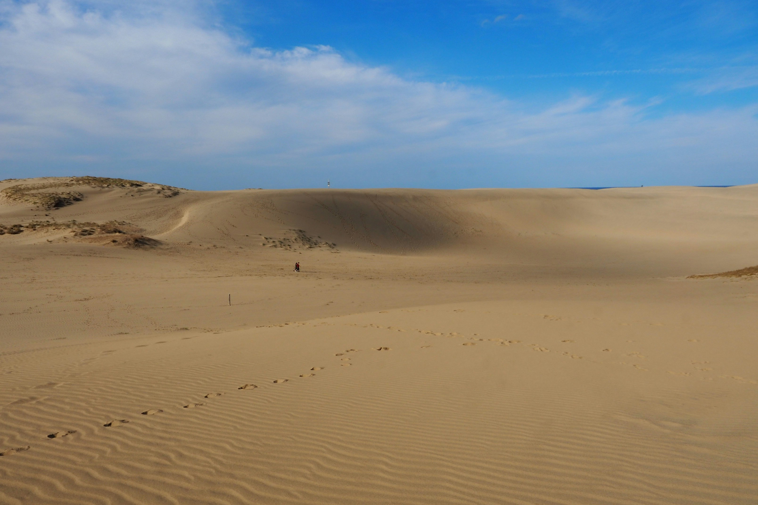 Expansive sandy dunes under a vibrant blue sky, marked by a few scattered footprints.