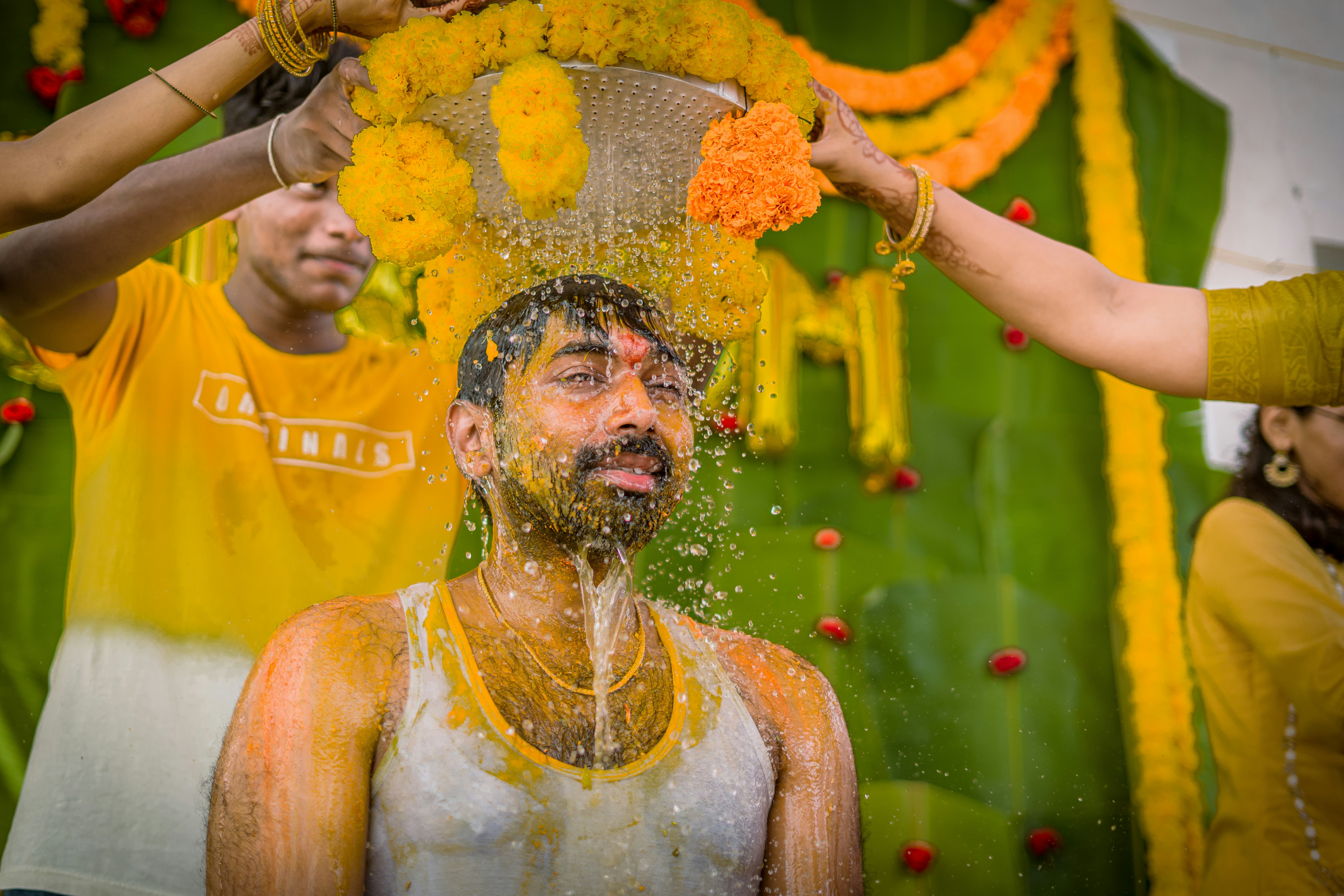 A man undergoing a ceremonial water blessing, surrounded by vibrant marigold flowers and festive decorations. The moment captures the essence of cultural celebration.