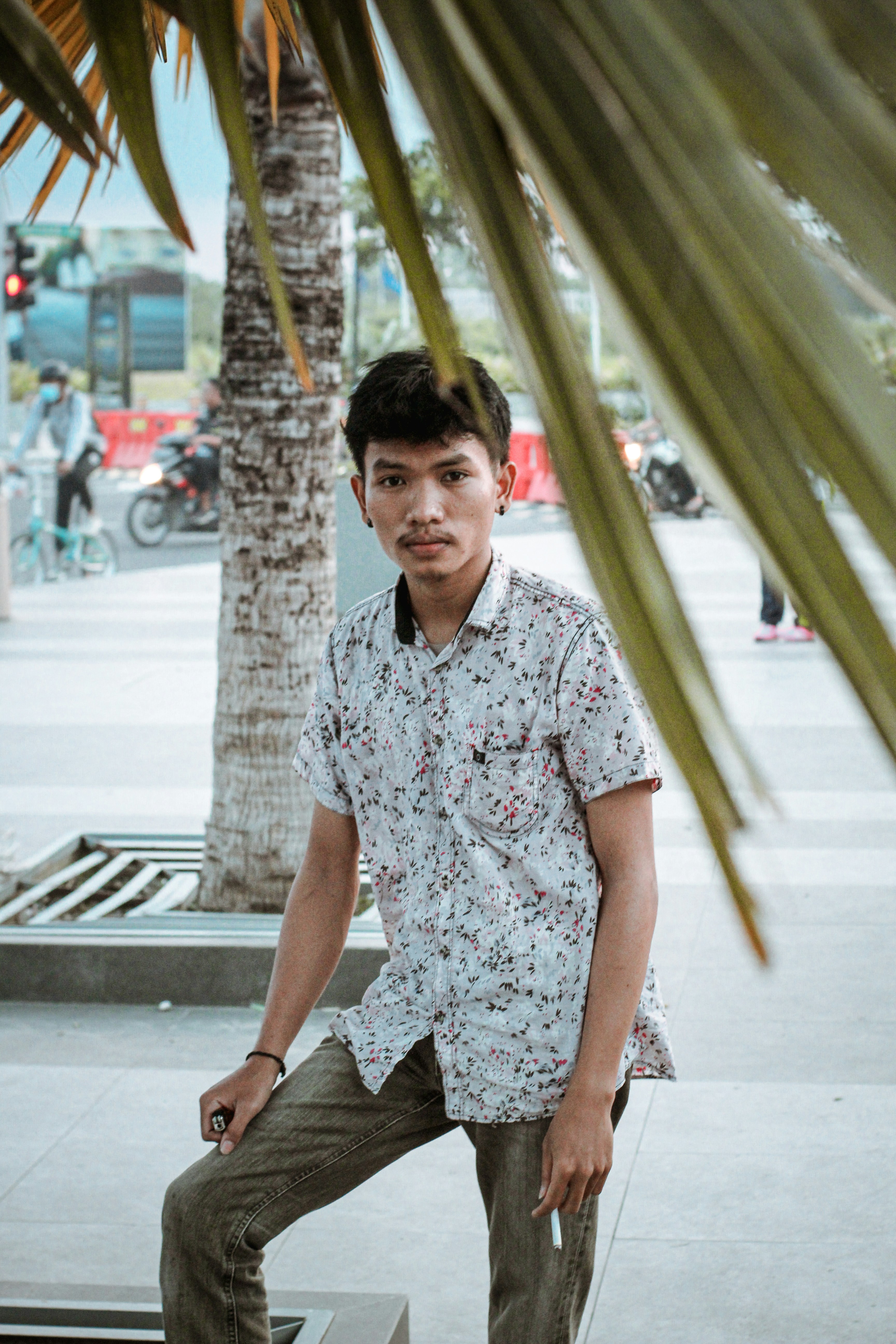 Young man seated casually in an urban setting, framed by palm leaves and bustling city life in the background.