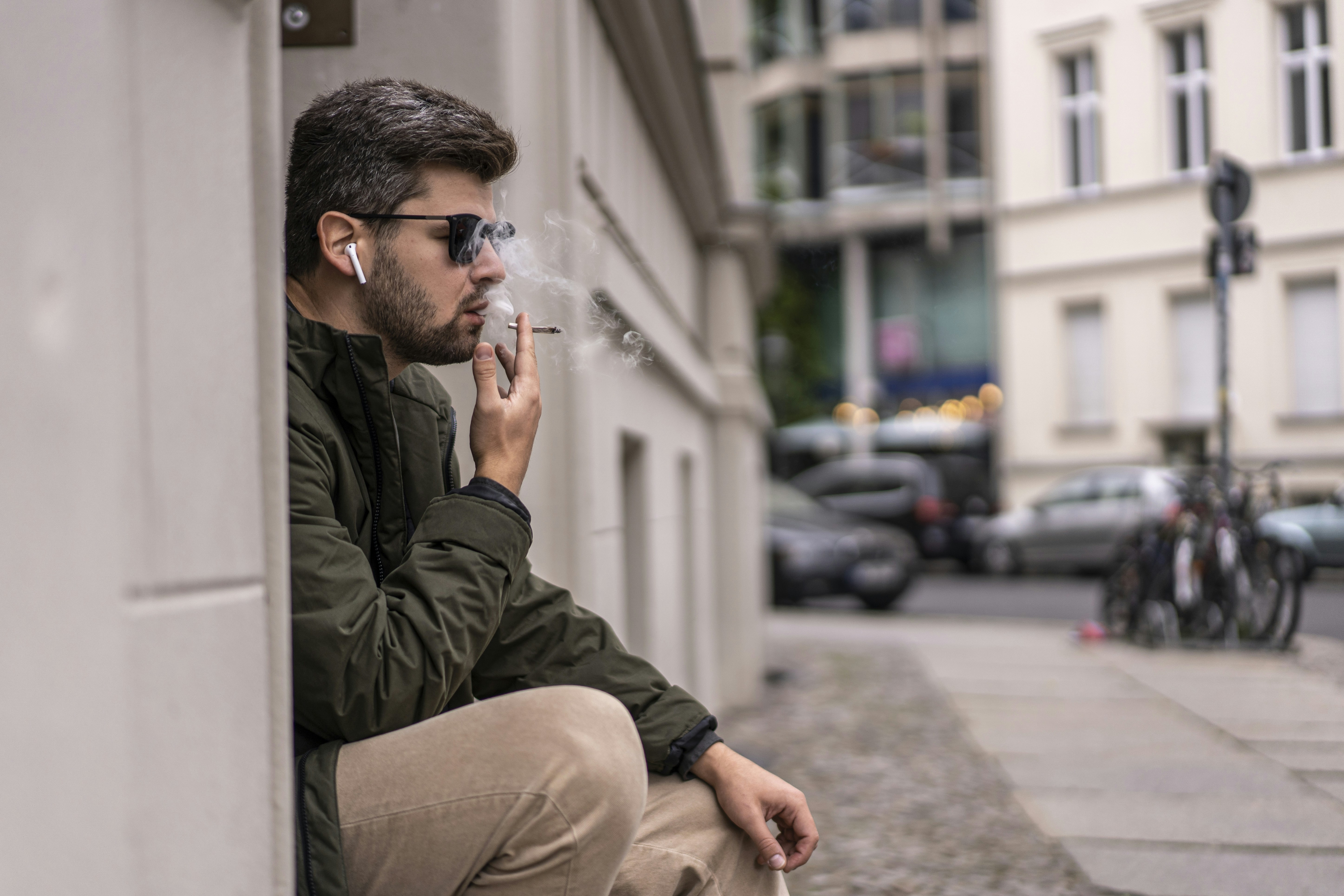 Man sitting against a wall, smoking a cigarette while wearing wireless earbuds in an urban setting.