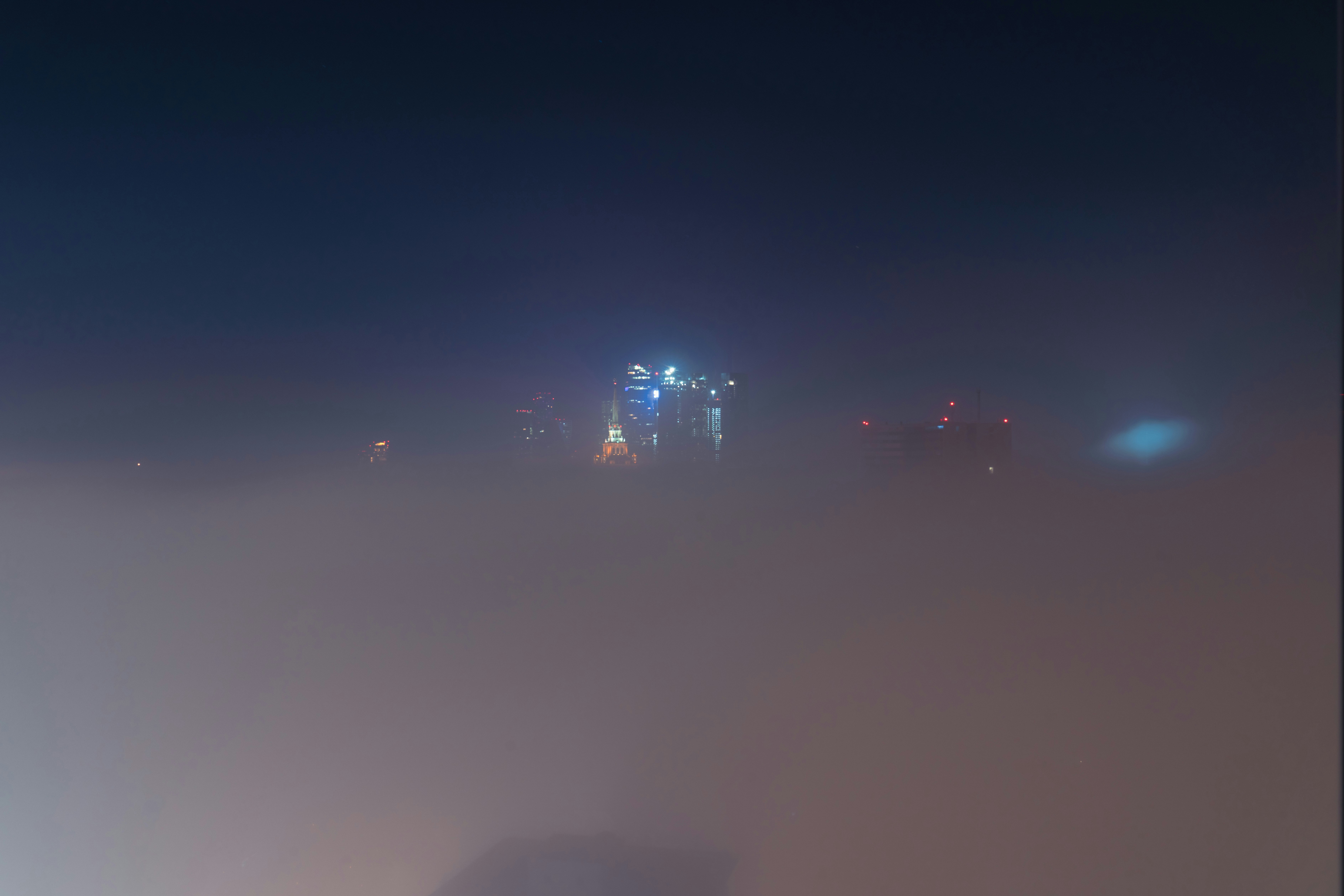 City skyline partially obscured by thick fog, with illuminated buildings peeking through the mist. A tranquil yet mysterious atmosphere envelops the scene.