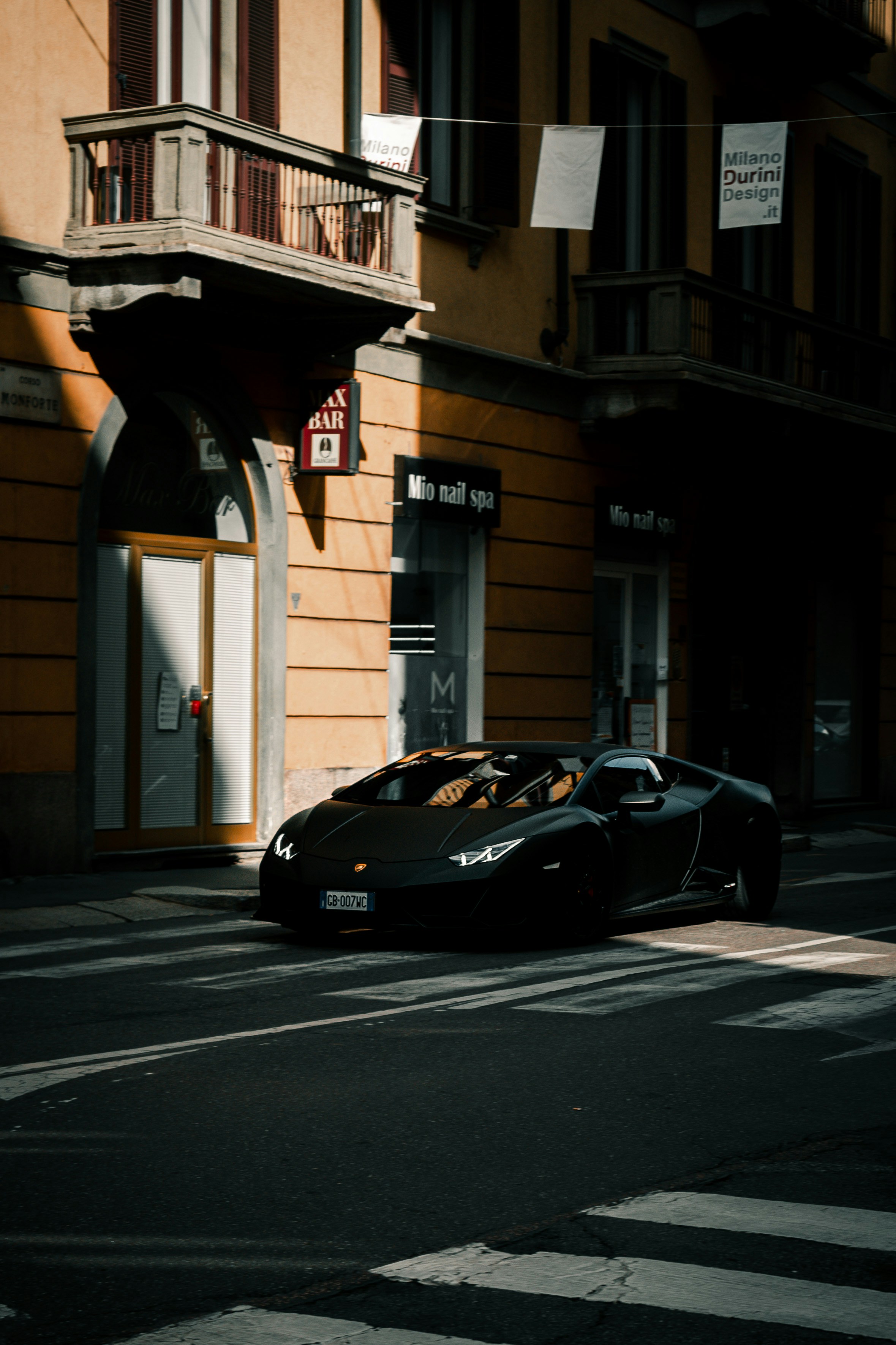 A sleek black Lamborghini parked on a city street, contrasting with the warm tones of the surrounding architecture. The interplay of light and shadow adds depth to the scene.