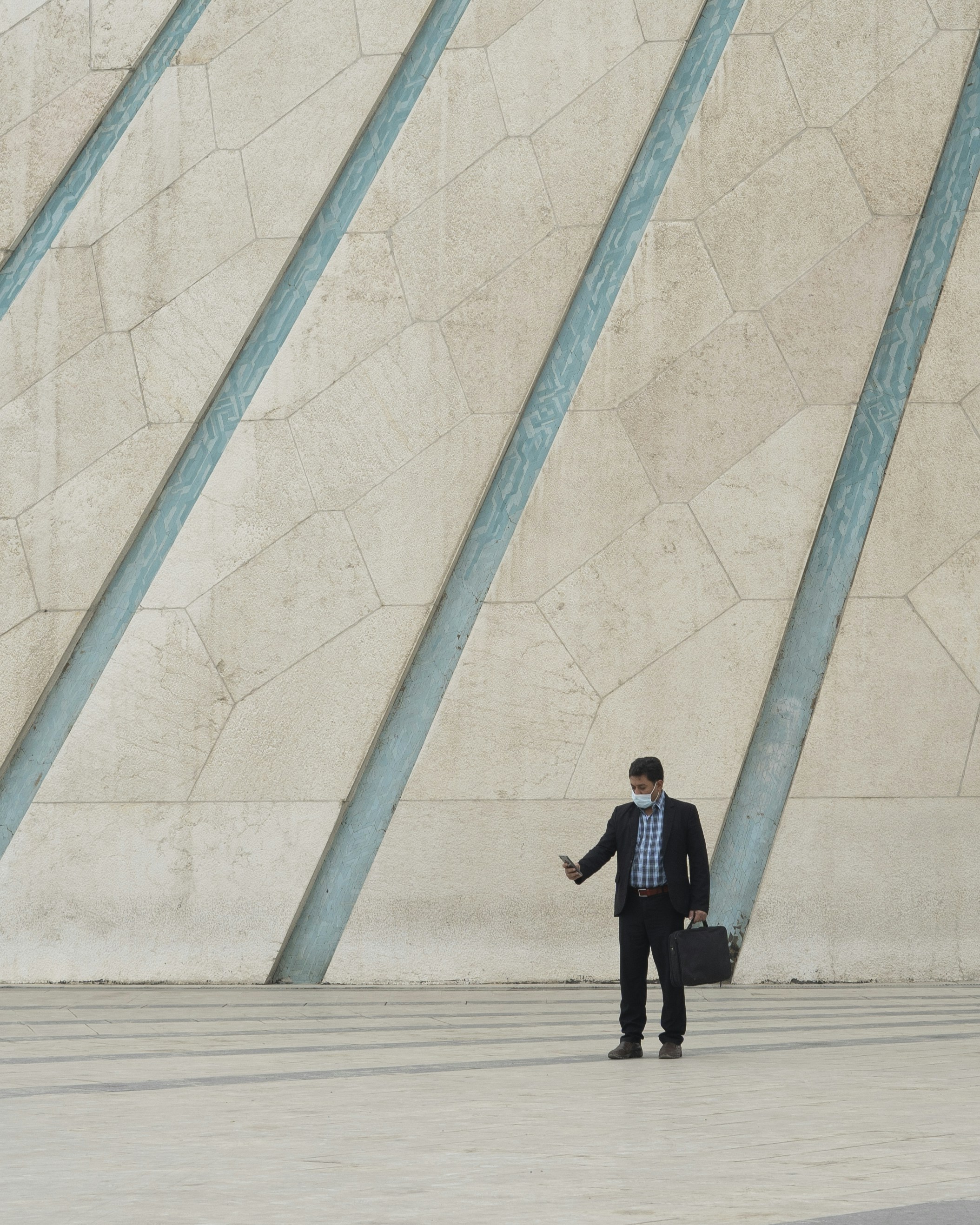 Man with briefcase and mask stands near diagonal architectural ribs in a modern plaza