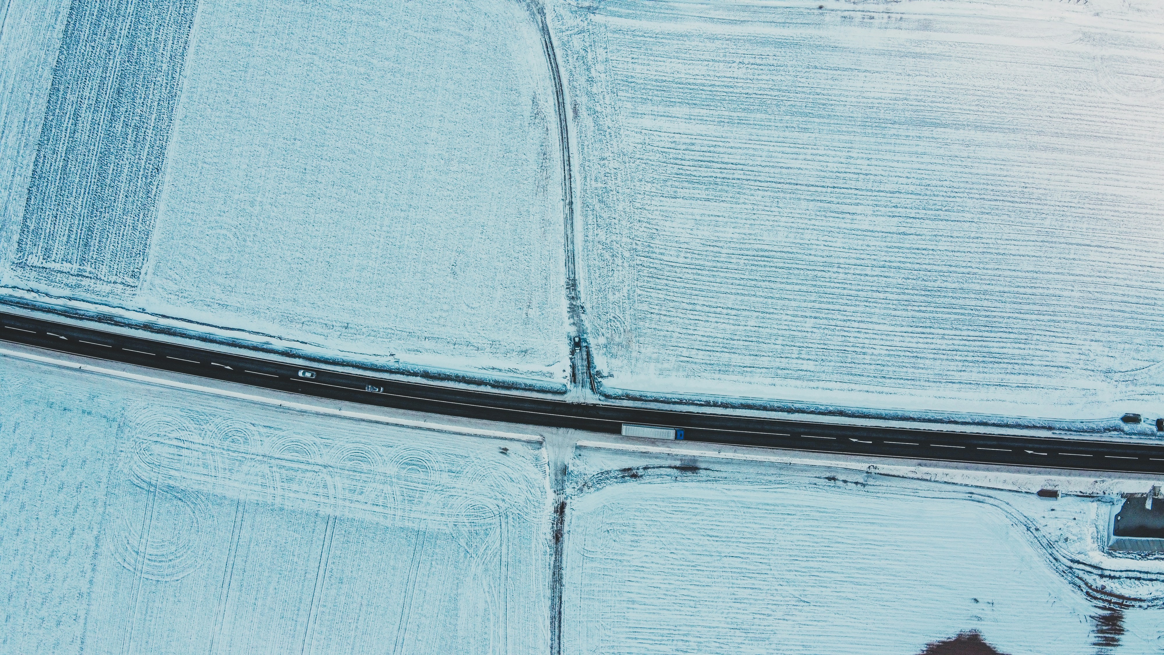Aerial view of a snow-covered road meandering through frost-laden fields, showcasing the contrast between the dark asphalt and the white terrain.