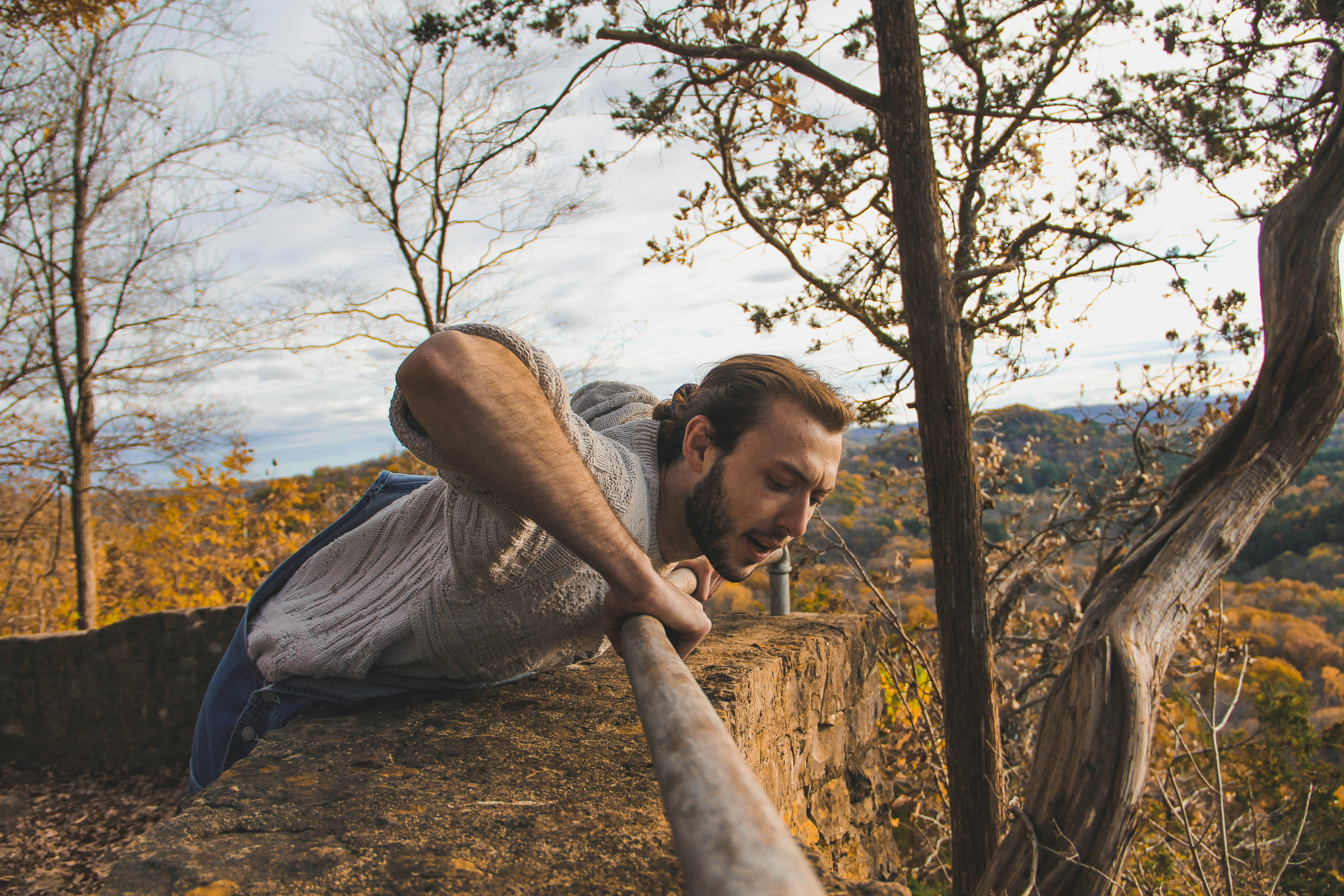 A man leans over a stone ledge, gripping a railing with determination, surrounded by autumn foliage and a distant landscape.