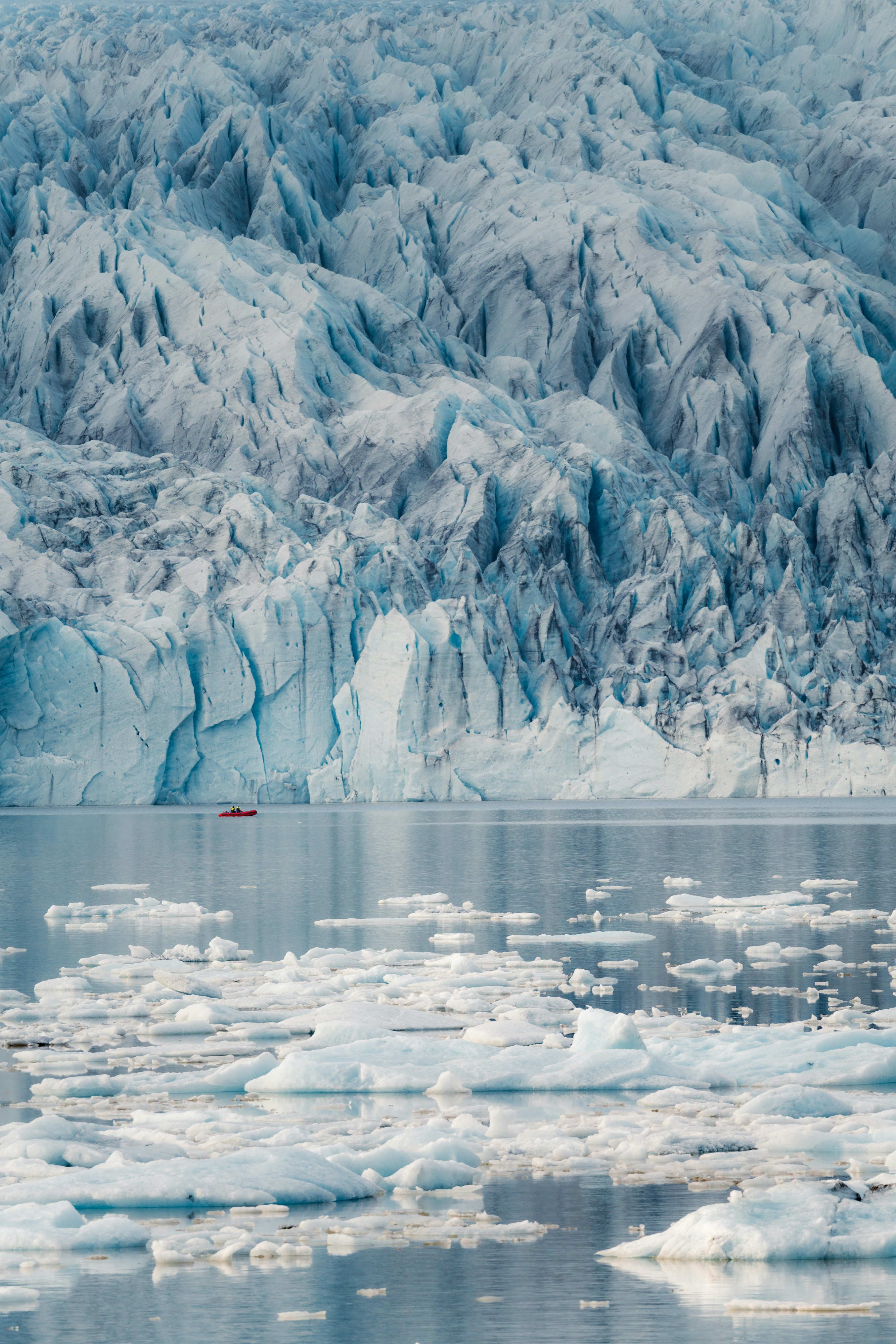 A vibrant red kayak navigates through a serene glacial landscape, surrounded by floating icebergs and towering icy cliffs. The scene captures the stark contrast between the vivid kayak and the cool tones of the glacier.