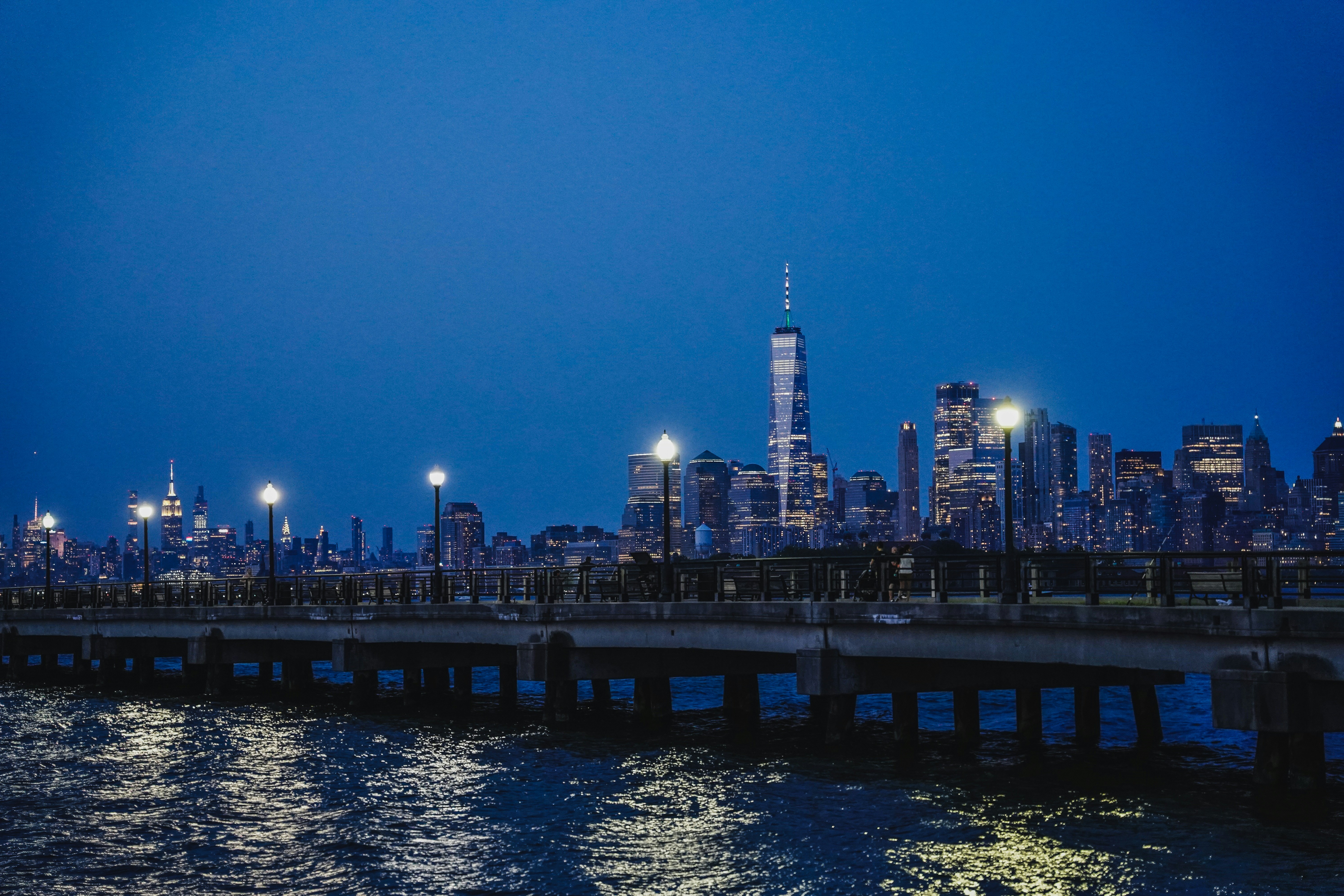 Manhattan skyline illuminated against a deep blue twilight sky, with lights reflecting on the water in the foreground.