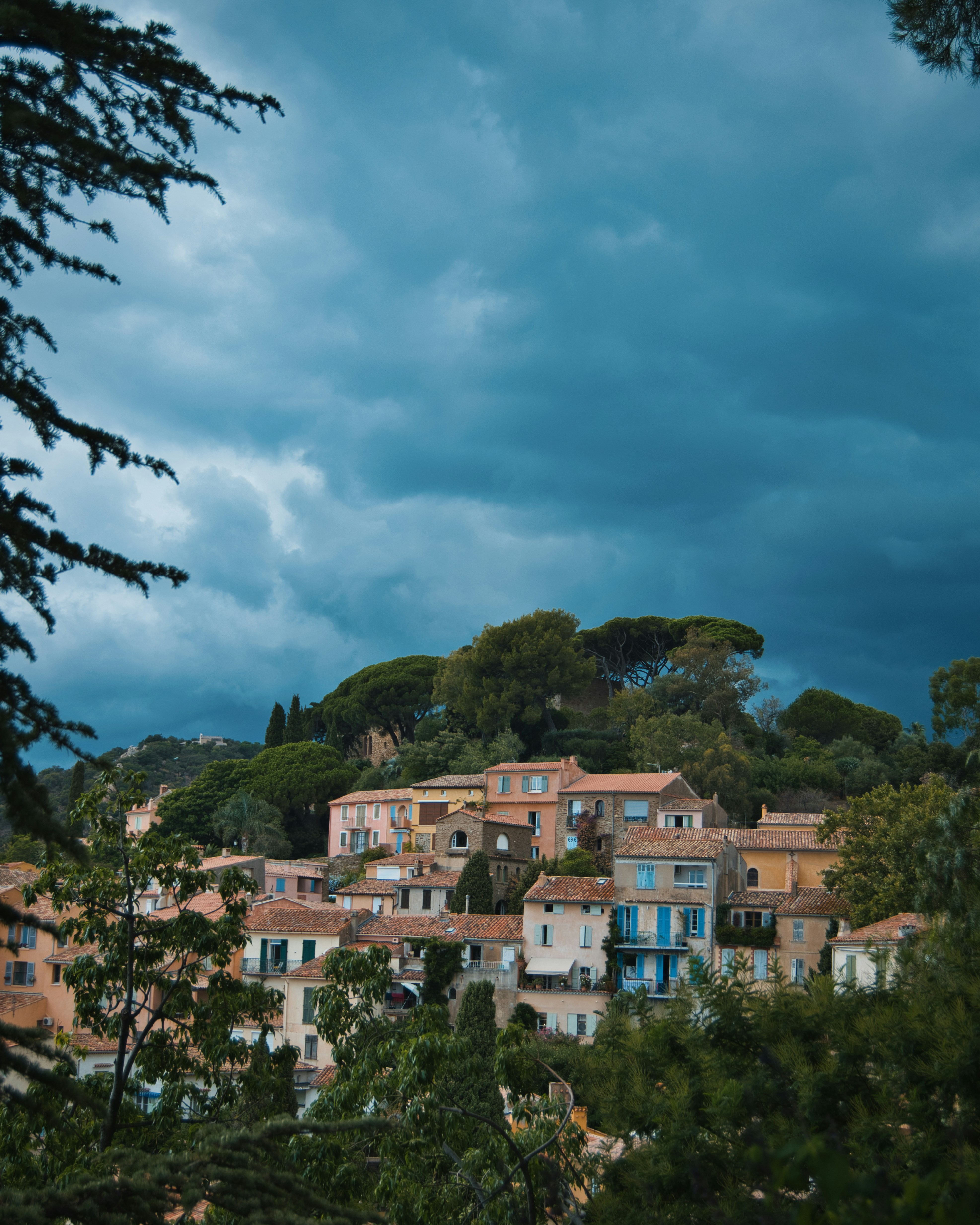 Charming hillside village nestled among lush trees, with pastel-colored houses under dramatic storm clouds. The scene evokes a sense of tranquility and impending change.