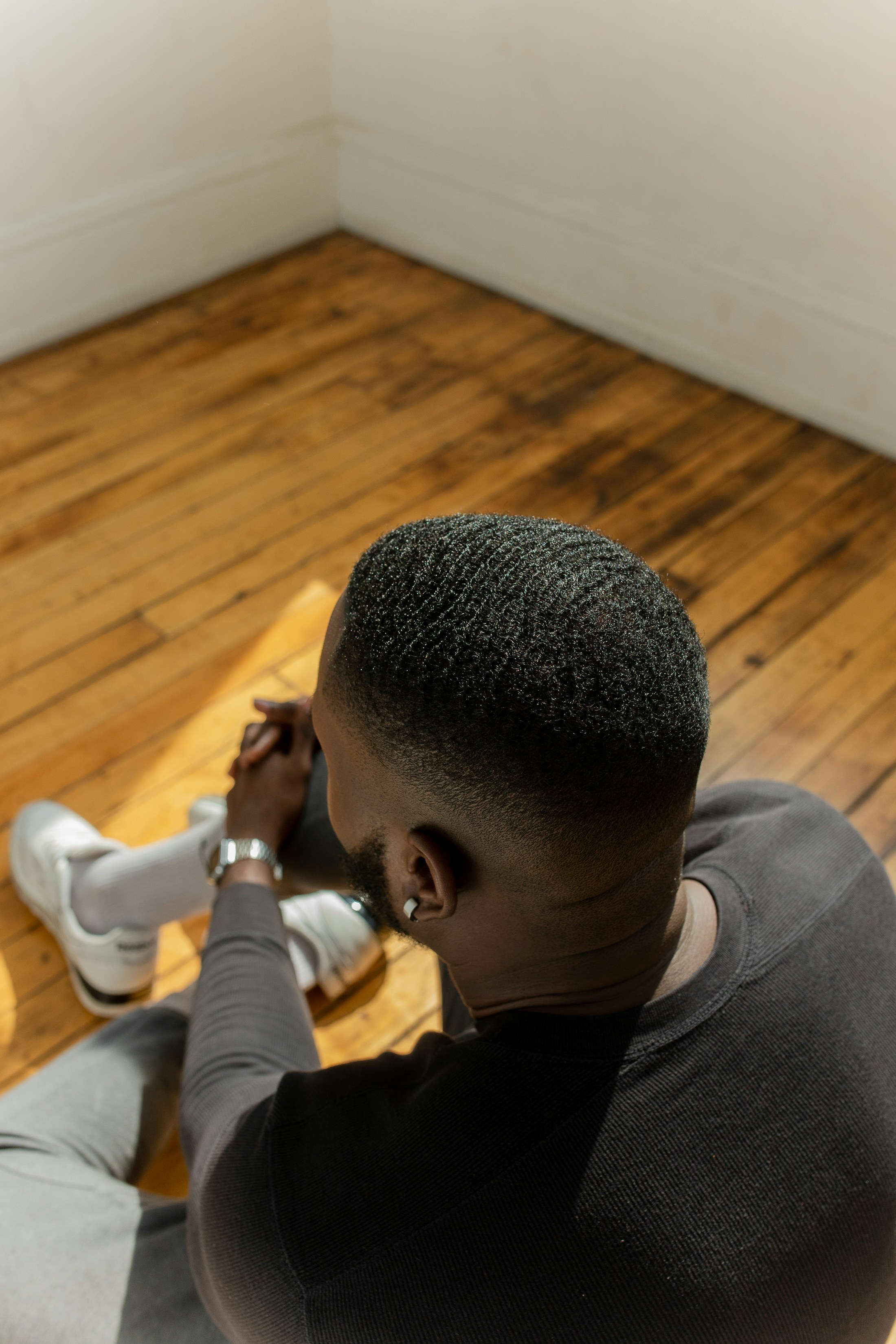 A man seated in a sunlit room, deep in thought, with a focus on his textured hairstyle and relaxed posture. The warm wooden floor adds to the tranquil atmosphere.