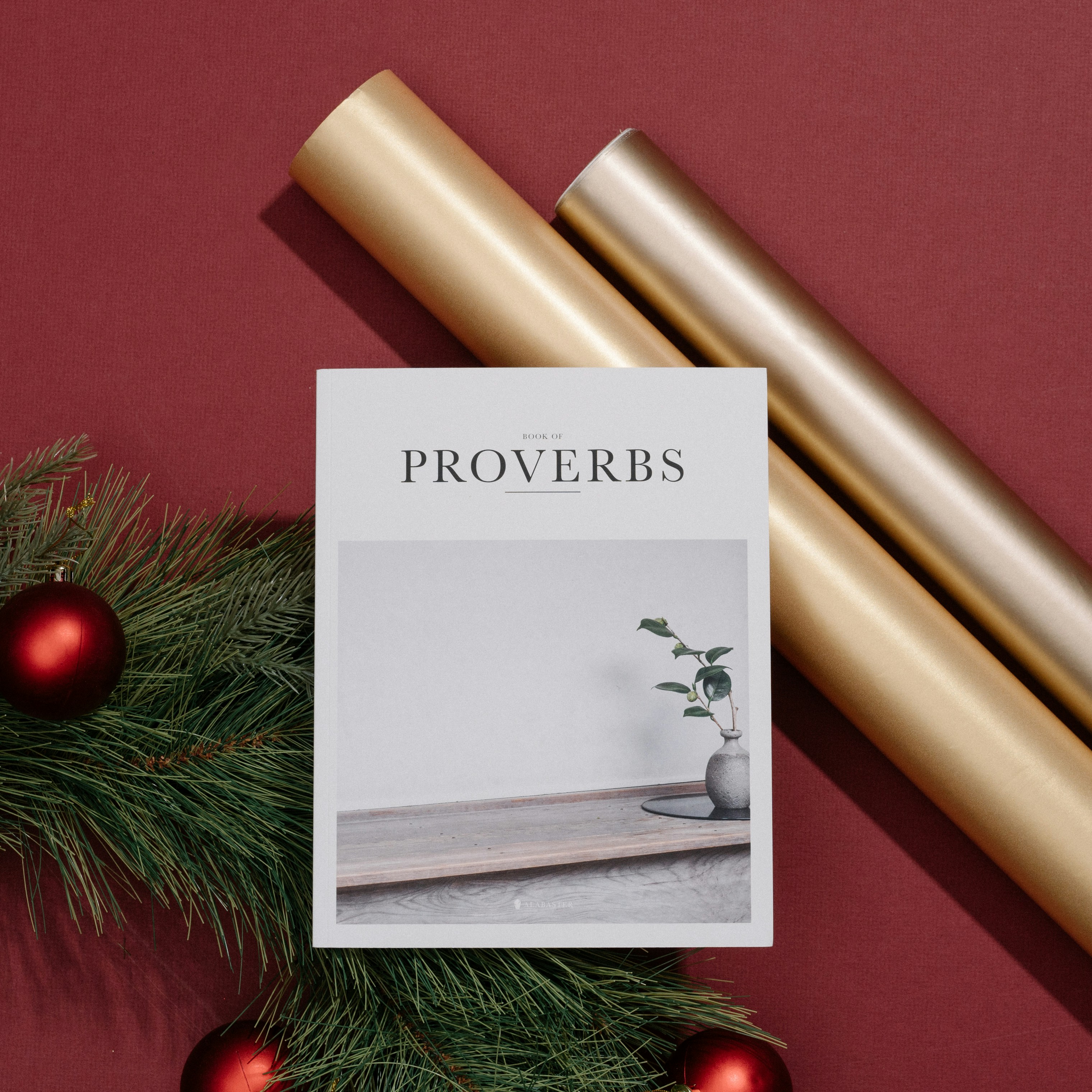 A minimalist book titled 'PROVERBS' rests on a wooden surface, surrounded by festive greenery and shiny gold wrapping paper rolls.