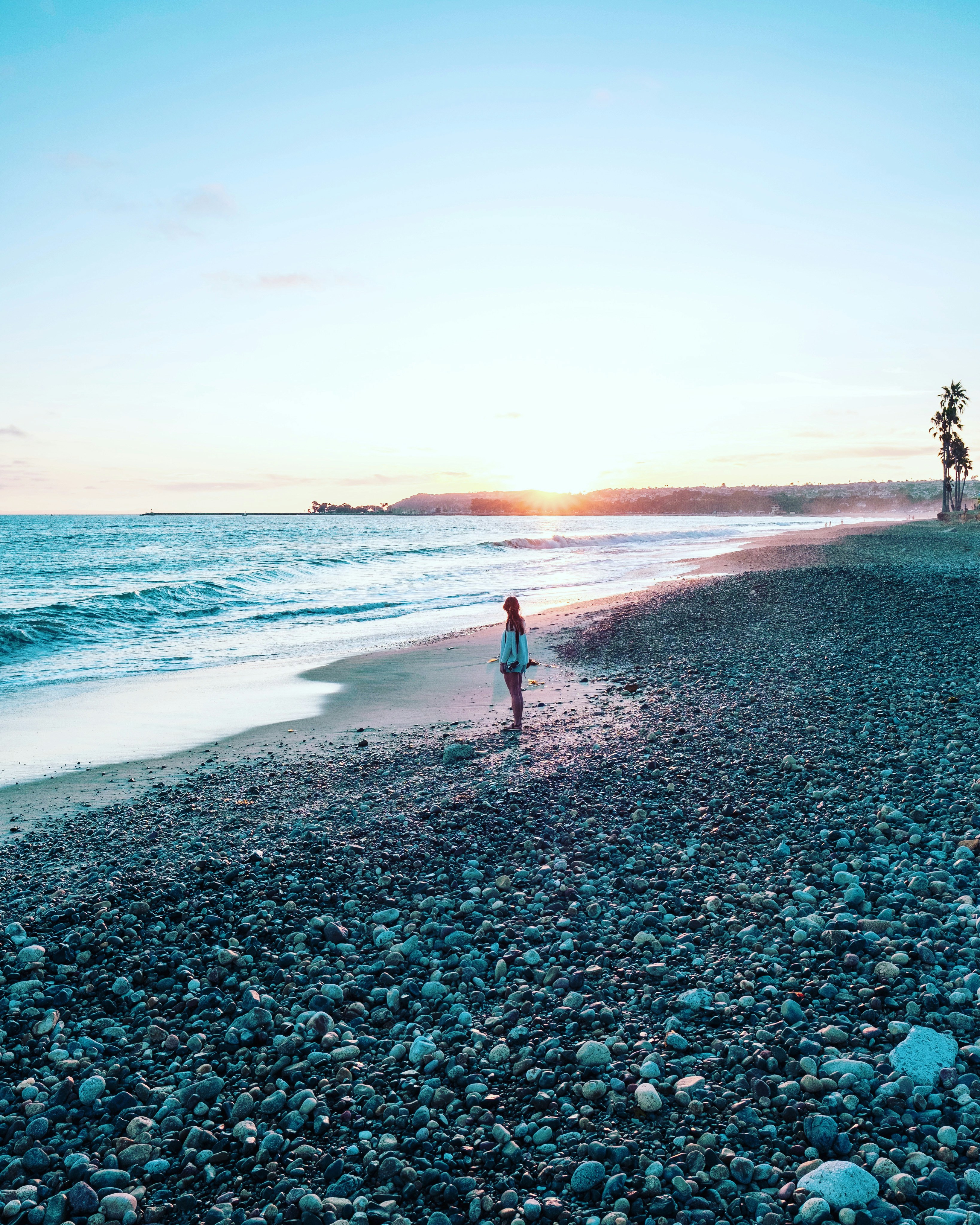 A solitary figure stands on a rocky beach at sunset, gazing at the tranquil waves and distant horizon. The scene captures the serene beauty of nature's transition from day to night.