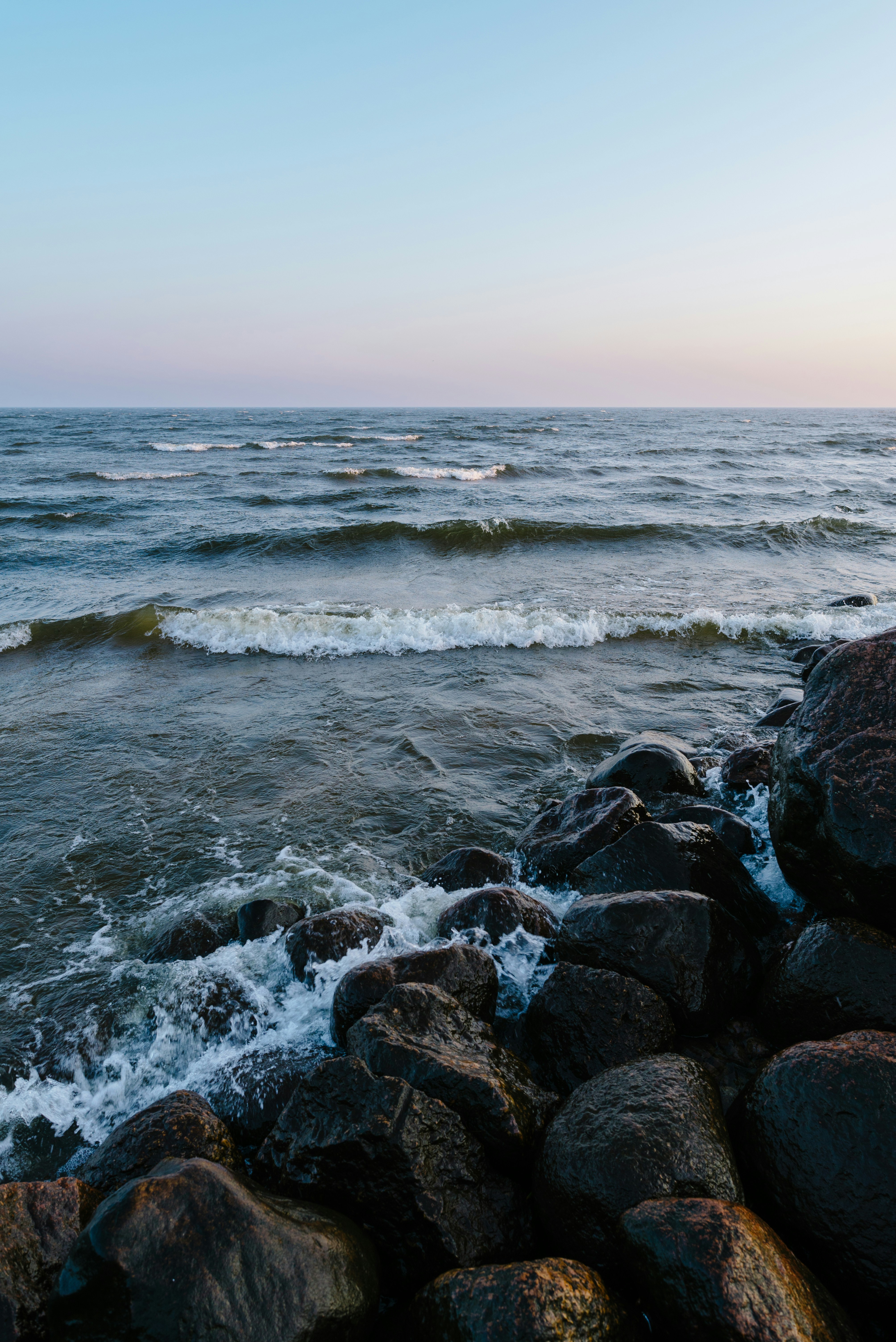 Gentle waves lap against rugged rocks as the day transitions to night, with a serene horizon blending soft hues. 