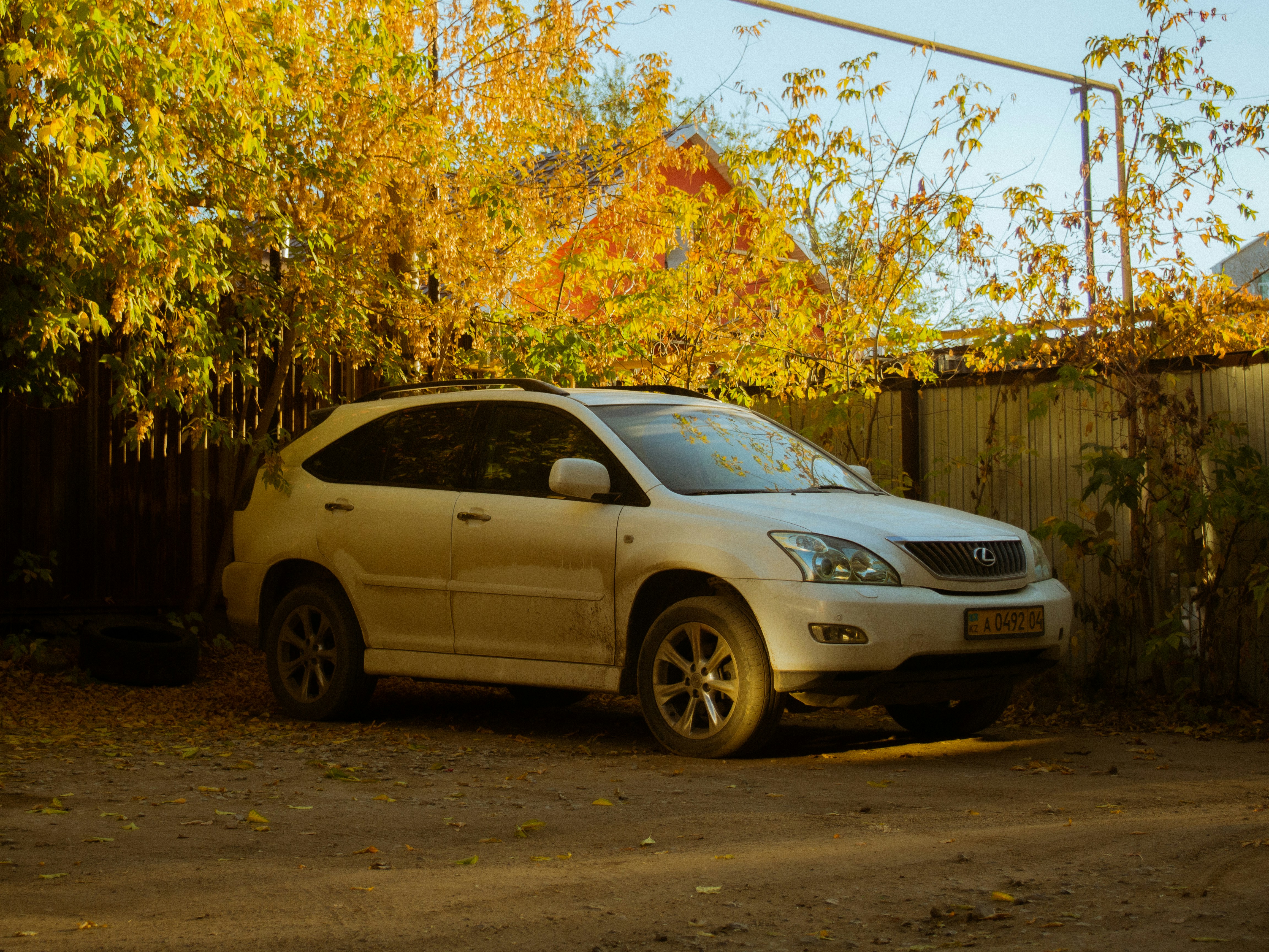A white SUV parked in a sunlit area surrounded by vibrant autumn leaves and a rustic fence.