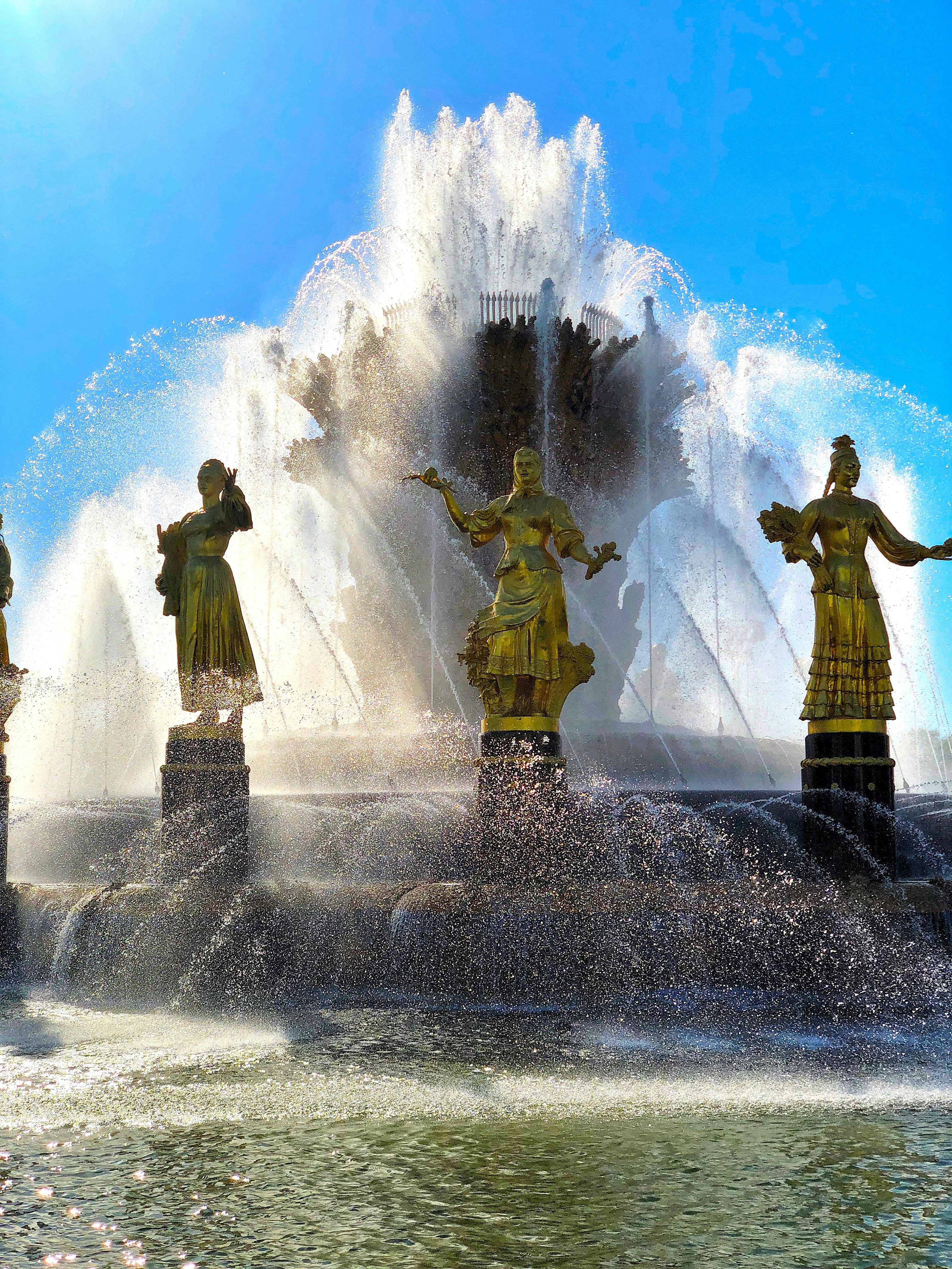 Golden statues gracefully emerge from a fountain, surrounded by a cascade of sparkling water against a clear blue sky.
