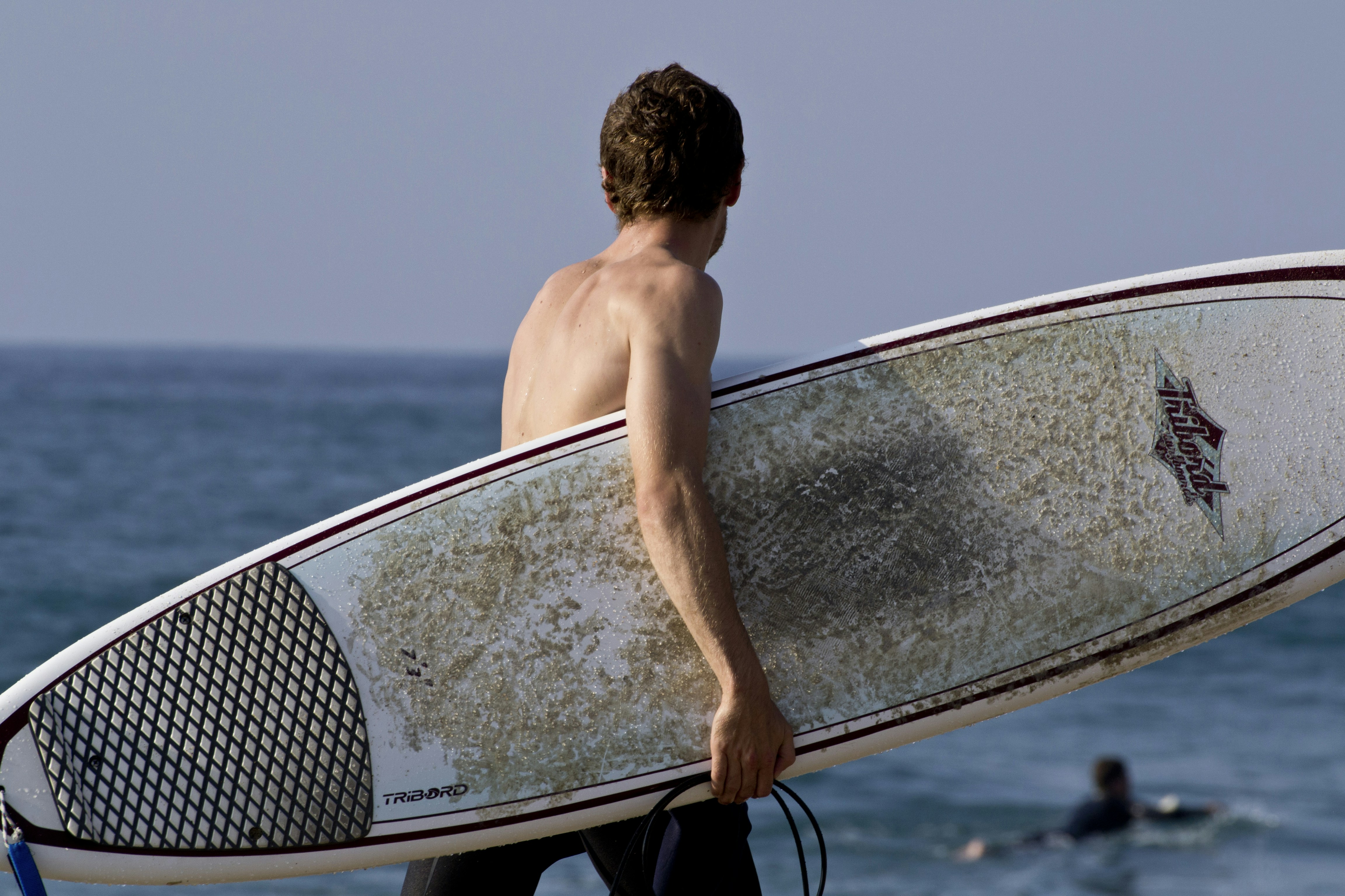 Surfer carrying a weathered board along the shoreline, with gentle waves in the background. The scene captures the anticipation of the surf ahead.