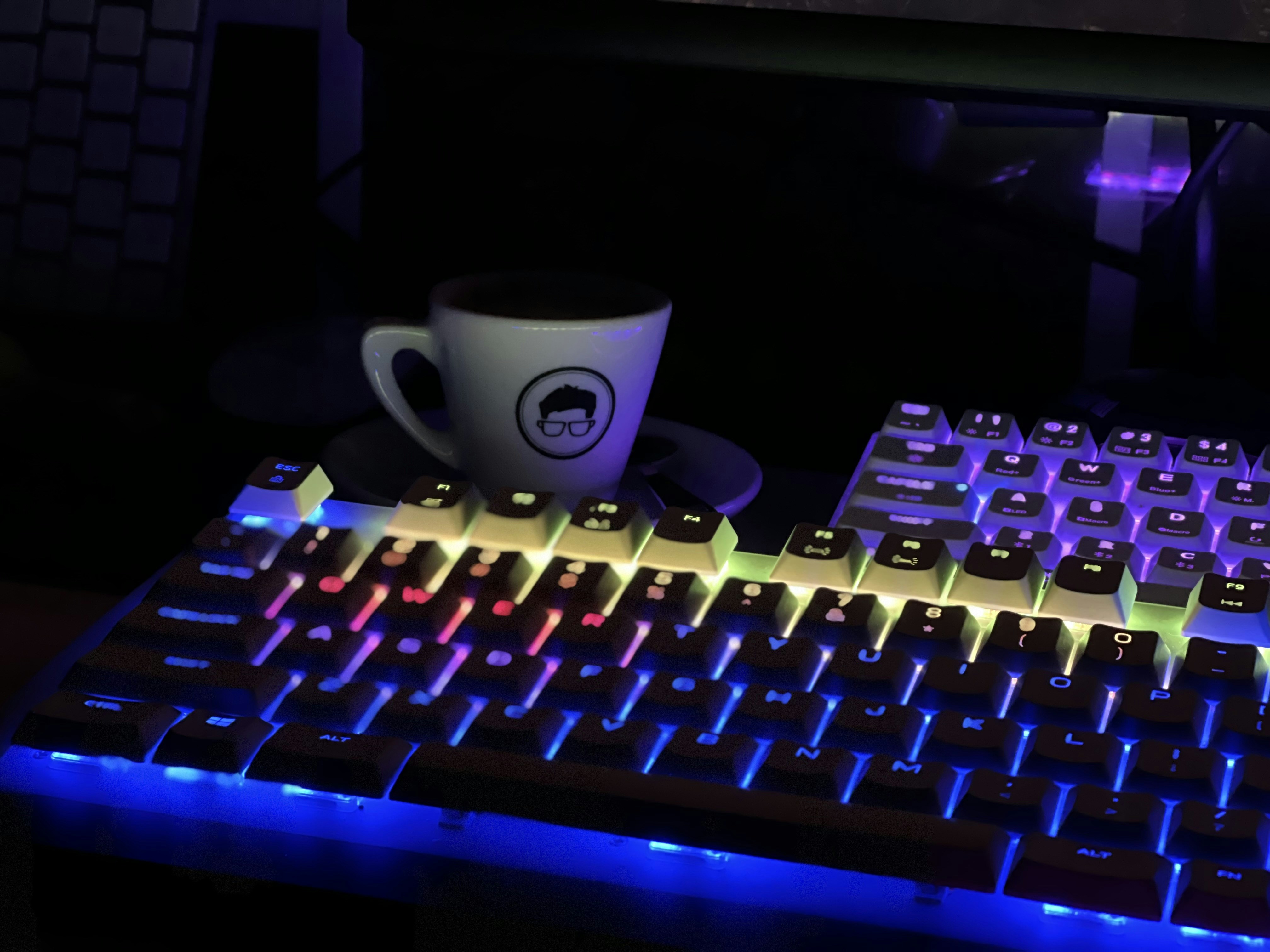 Vibrant gaming keyboard illuminated in blue and pink hues beside a small coffee cup with a playful design.