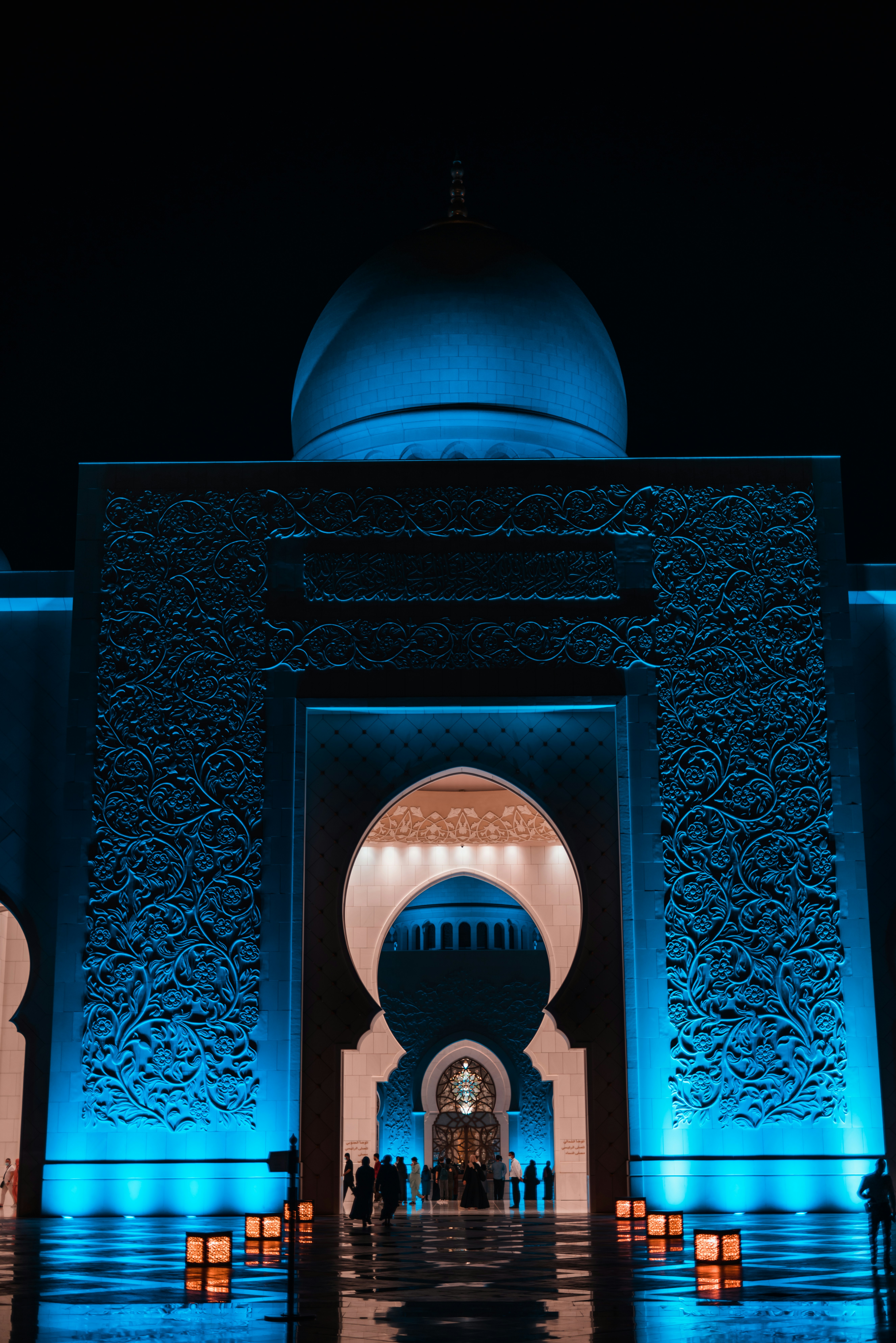 Intricate architectural details of a grand entrance illuminated in blue light, highlighting the artistry and design. Visitors gather at the threshold, creating a serene atmosphere.