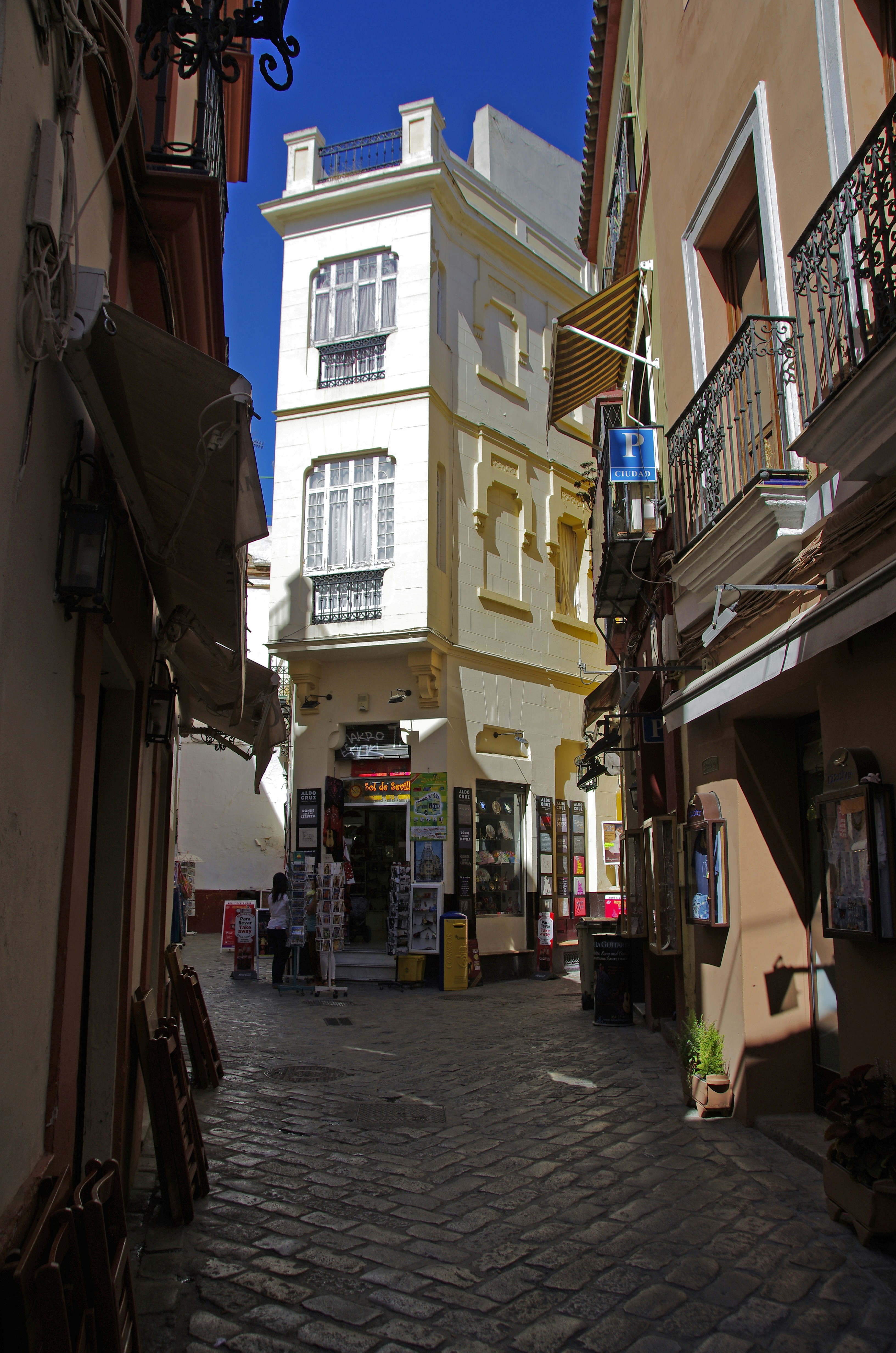 Charming narrow alleyway with a yellow building and a shop entrance, framed by cobblestones and colorful awnings. Bright blue sky peeks above the rooftops.