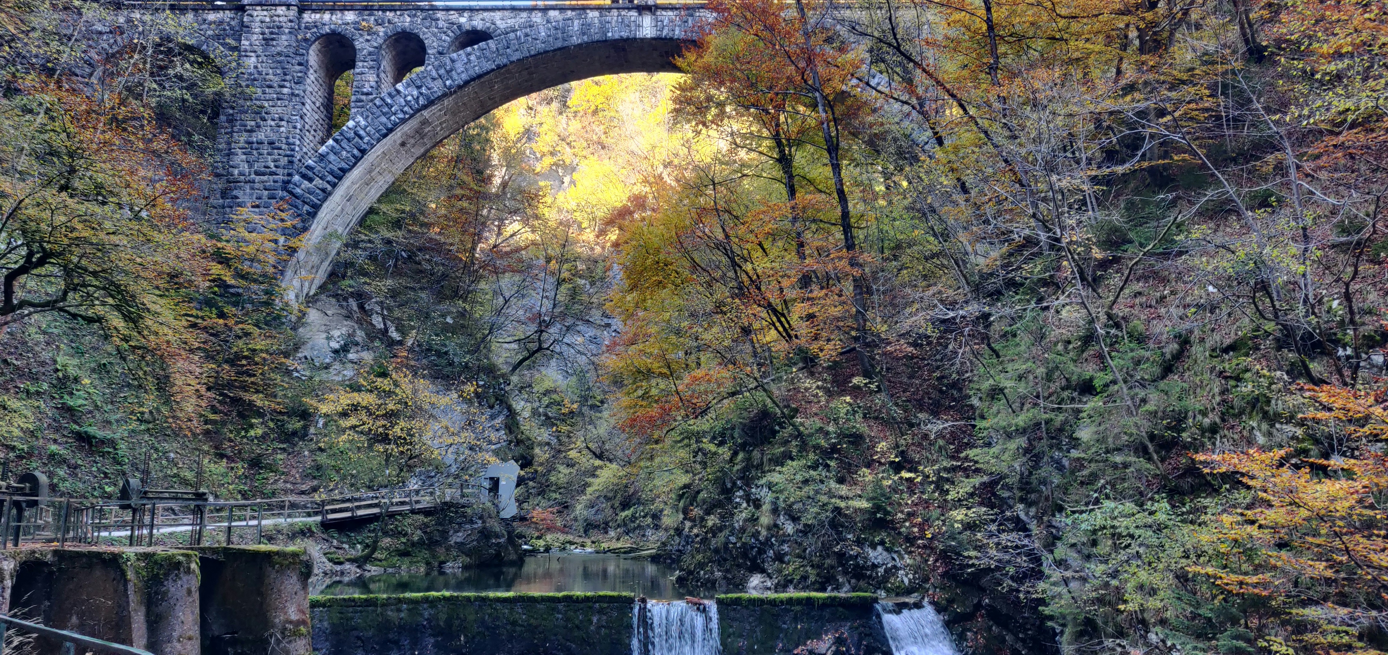 Historic stone bridge spanning a tranquil river, surrounded by vibrant autumn foliage and cascading waterfalls.