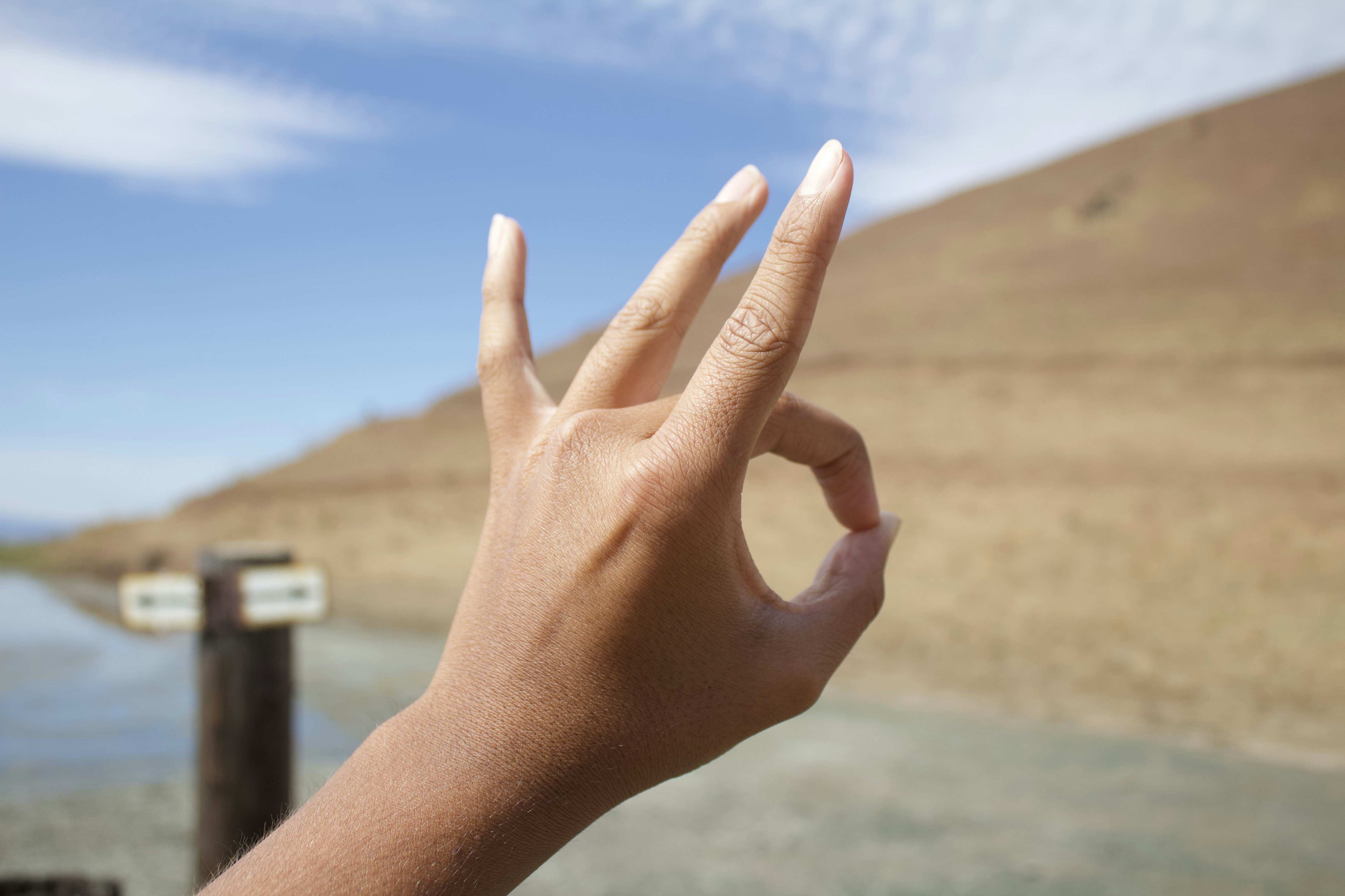 Hand forming an 'OK' gesture against a backdrop of a serene landscape with hills and water.
