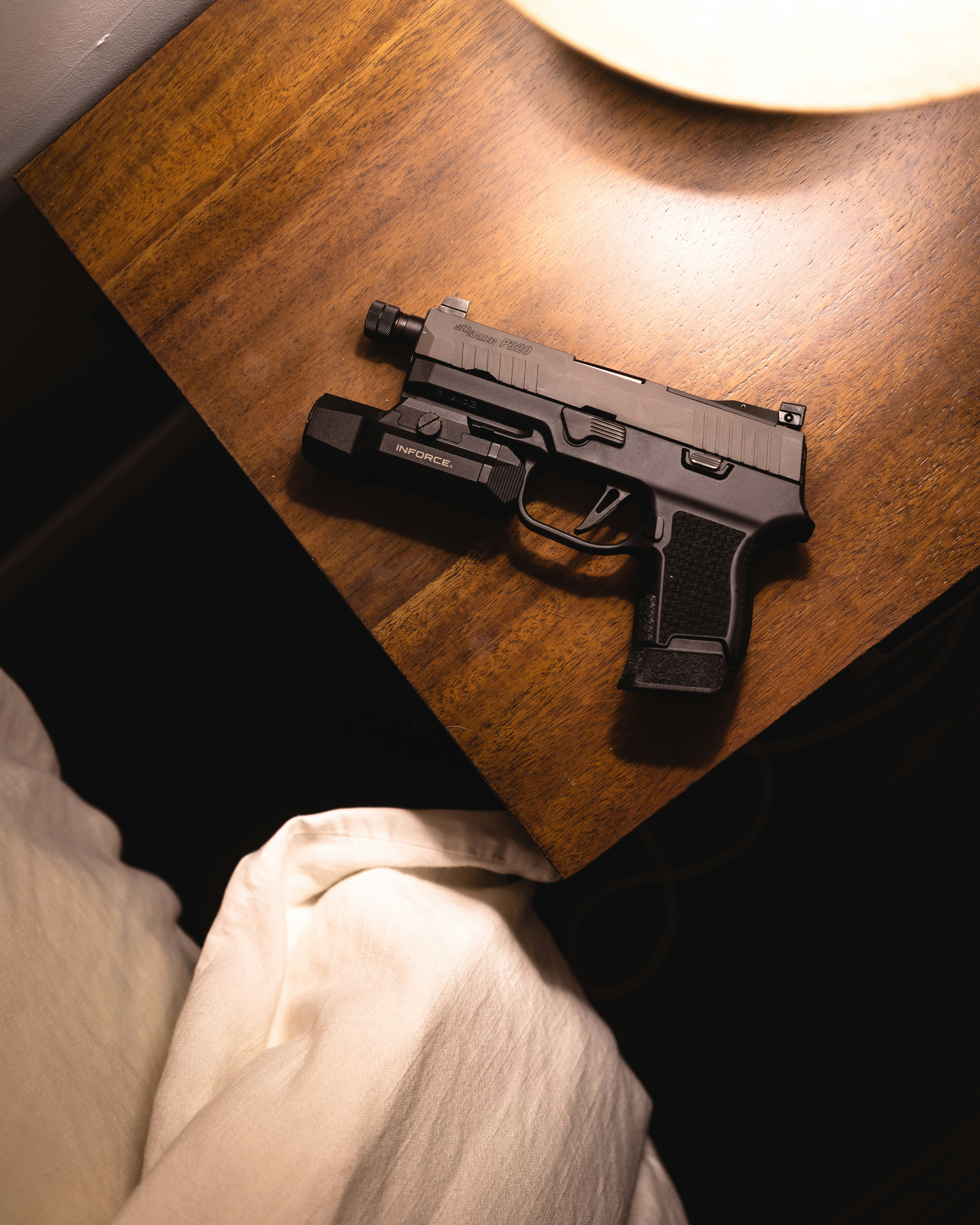 Handgun resting on a wooden nightstand, illuminated by soft light. A white fabric drapes subtly in the foreground.