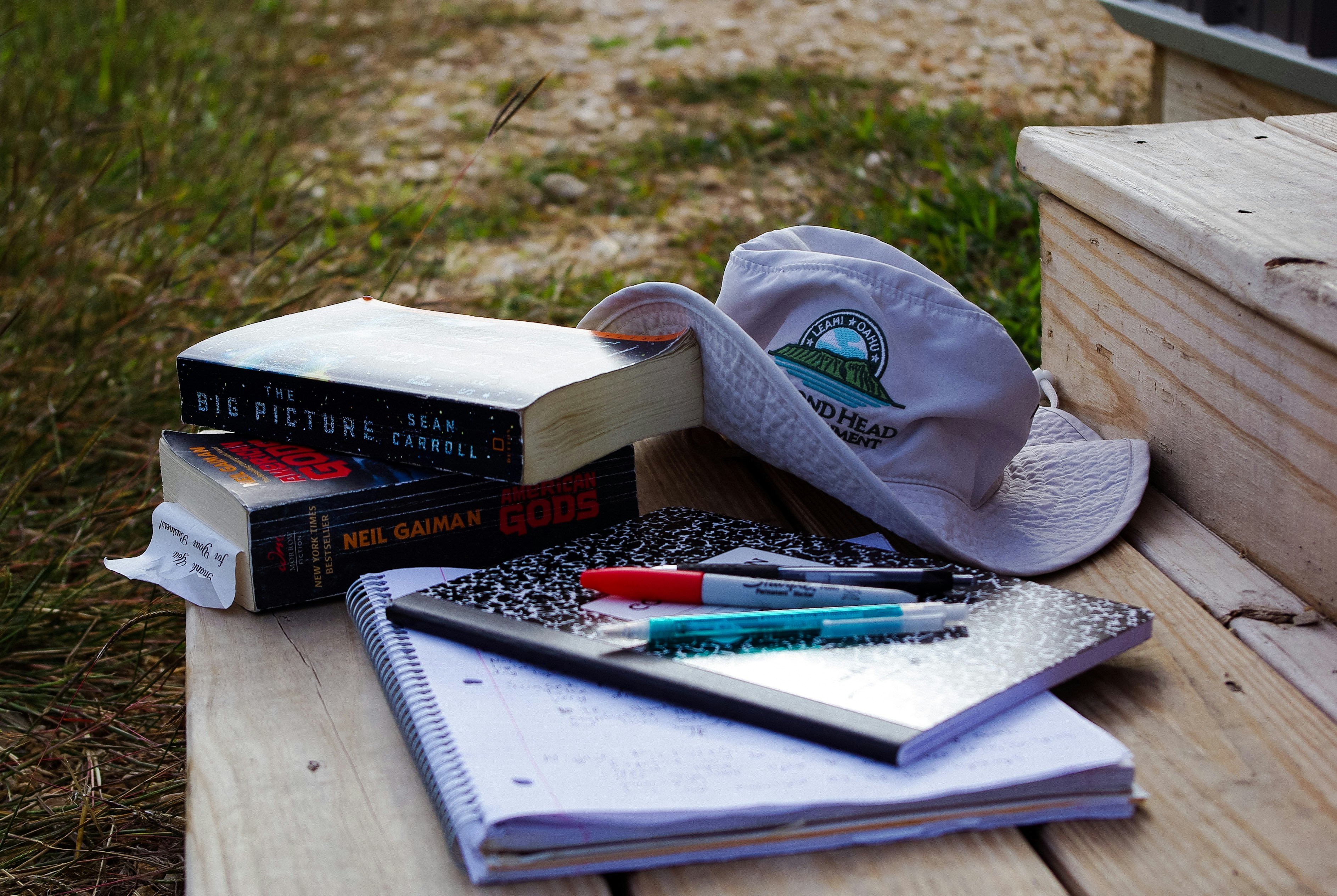 Stack of books alongside a notebook, pens, and a sun hat on wooden steps, suggesting a serene writing environment.