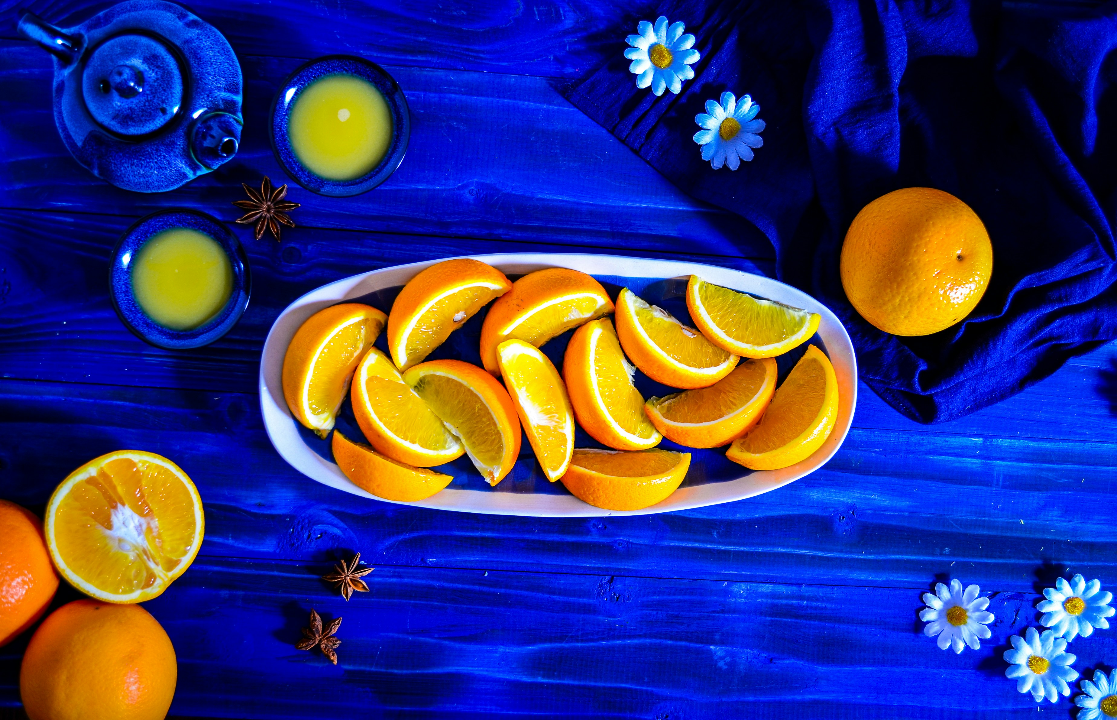 Sliced oranges arranged neatly on a white platter, surrounded by whole oranges, flowers, and a teapot on a vibrant blue background.