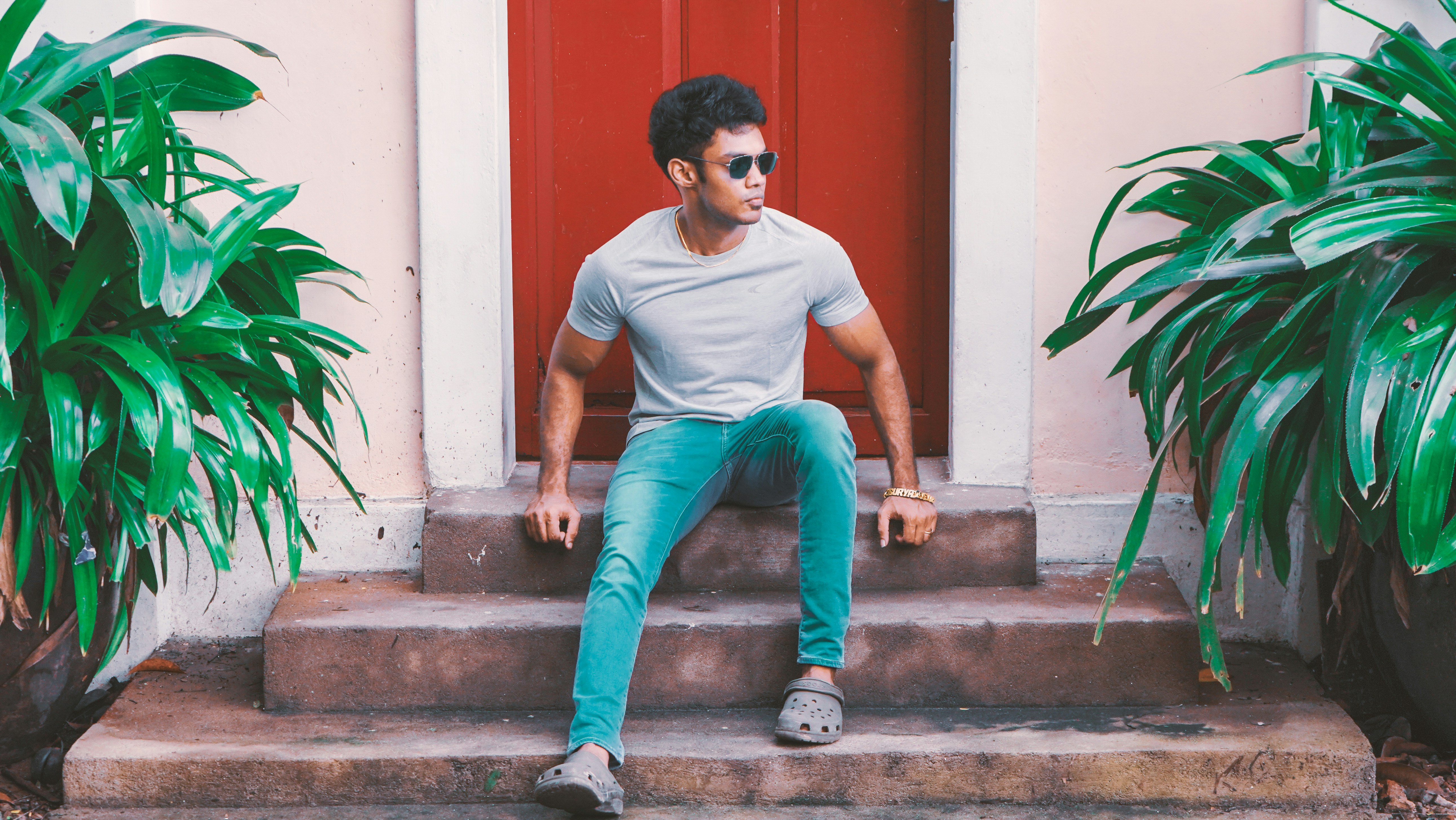 A young man seated on stone steps, flanked by lush green plants, gazes thoughtfully towards a vibrant red door.