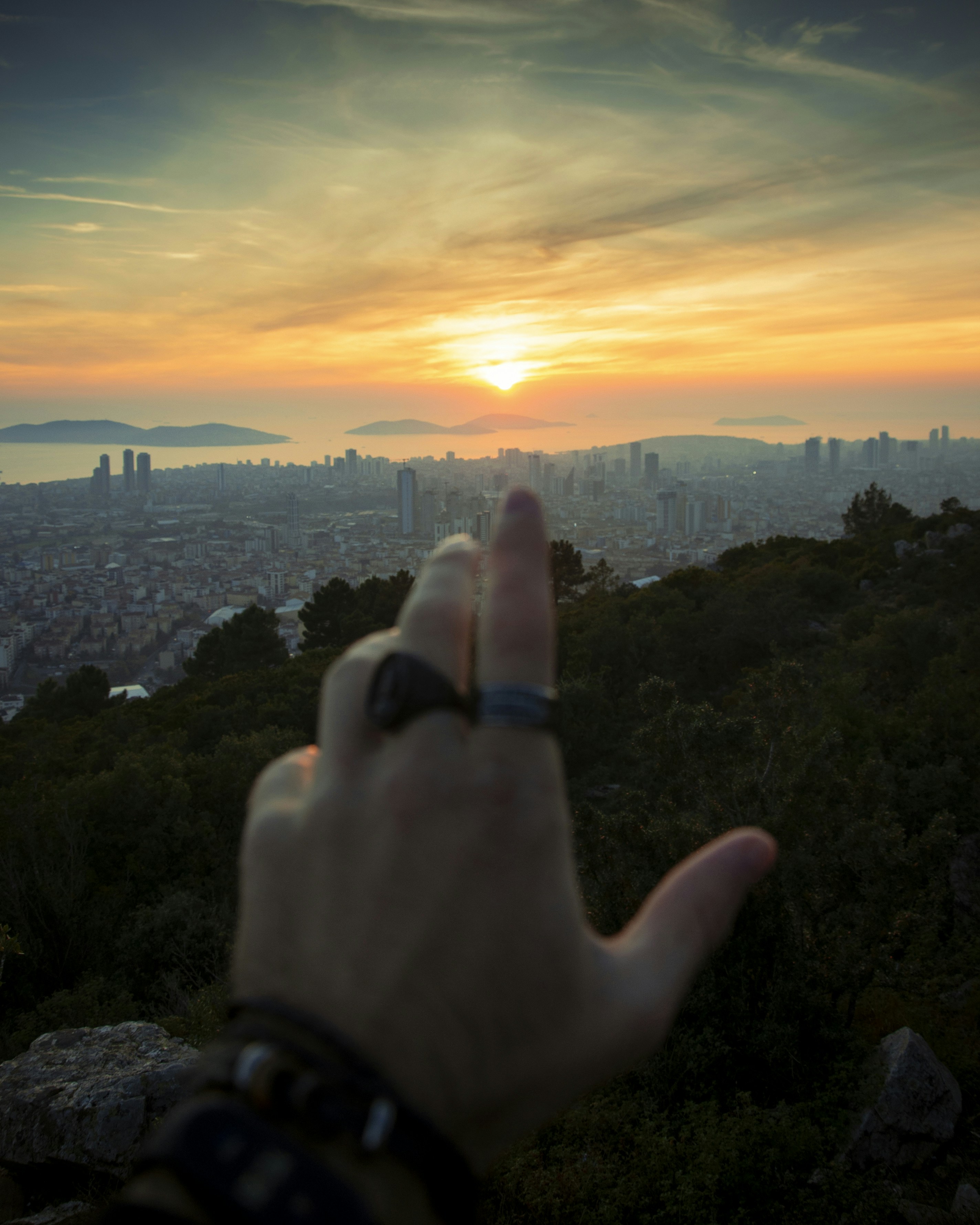 Outstretched hand reaching towards a vibrant sunset over a sprawling cityscape, with silhouettes of buildings and distant islands. 