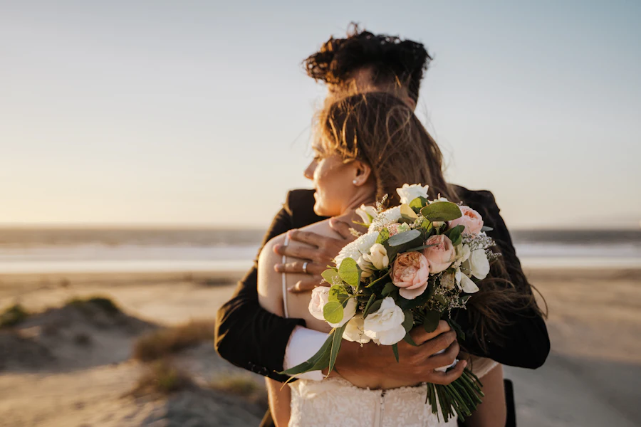 A bride and groom embrace on a beach at sunset, with the bride holding a bouquet of pale roses and greenery. The warm light and soft ocean background create a romantic scene.