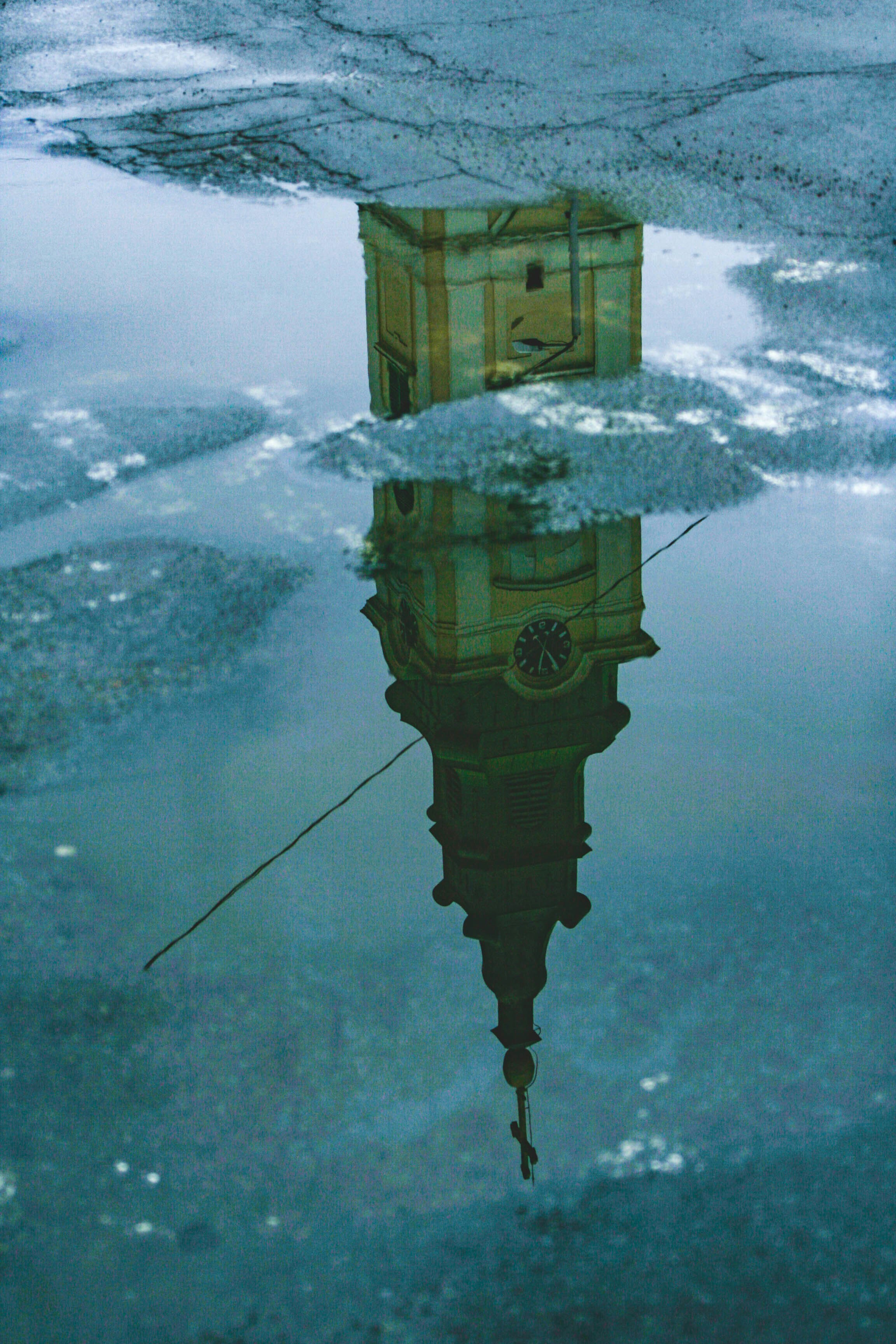 Clock tower reflected in a puddle, showcasing intricate architectural details and a moody atmosphere.
