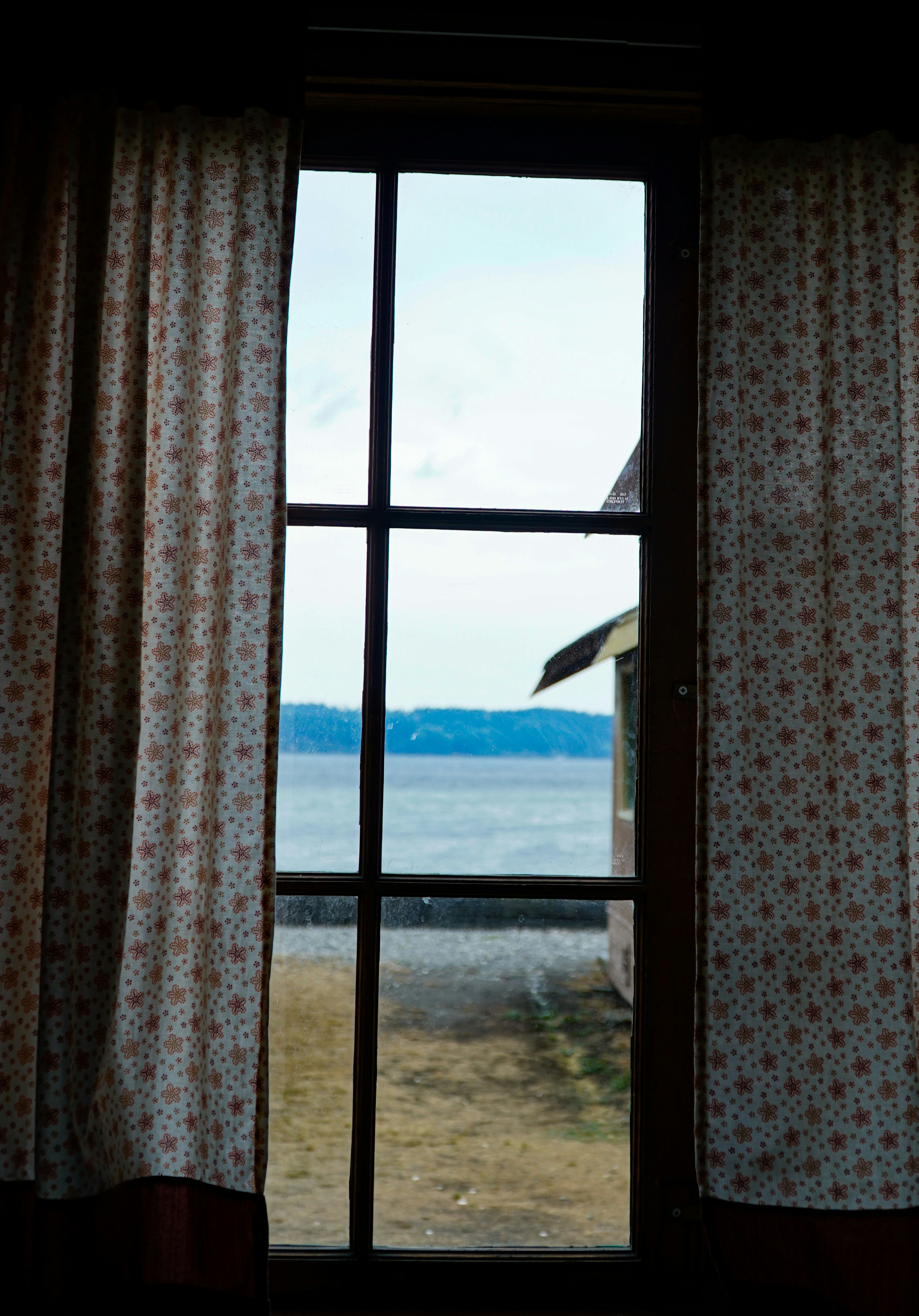 View through a window adorned with floral curtains, revealing a tranquil landscape of water and shoreline.