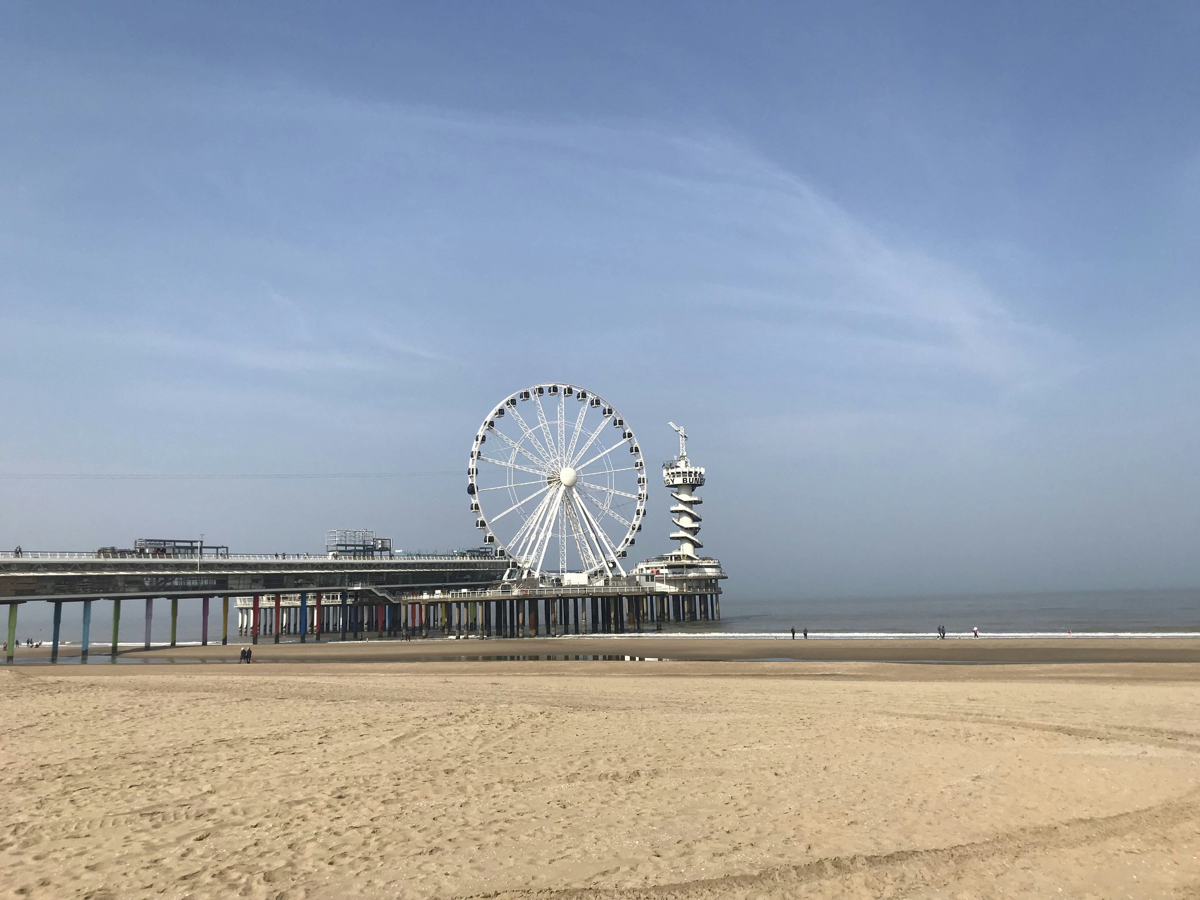 Ferris wheel towering over a bustling seaside pier against a clear blue sky. The sandy beach stretches in the foreground.