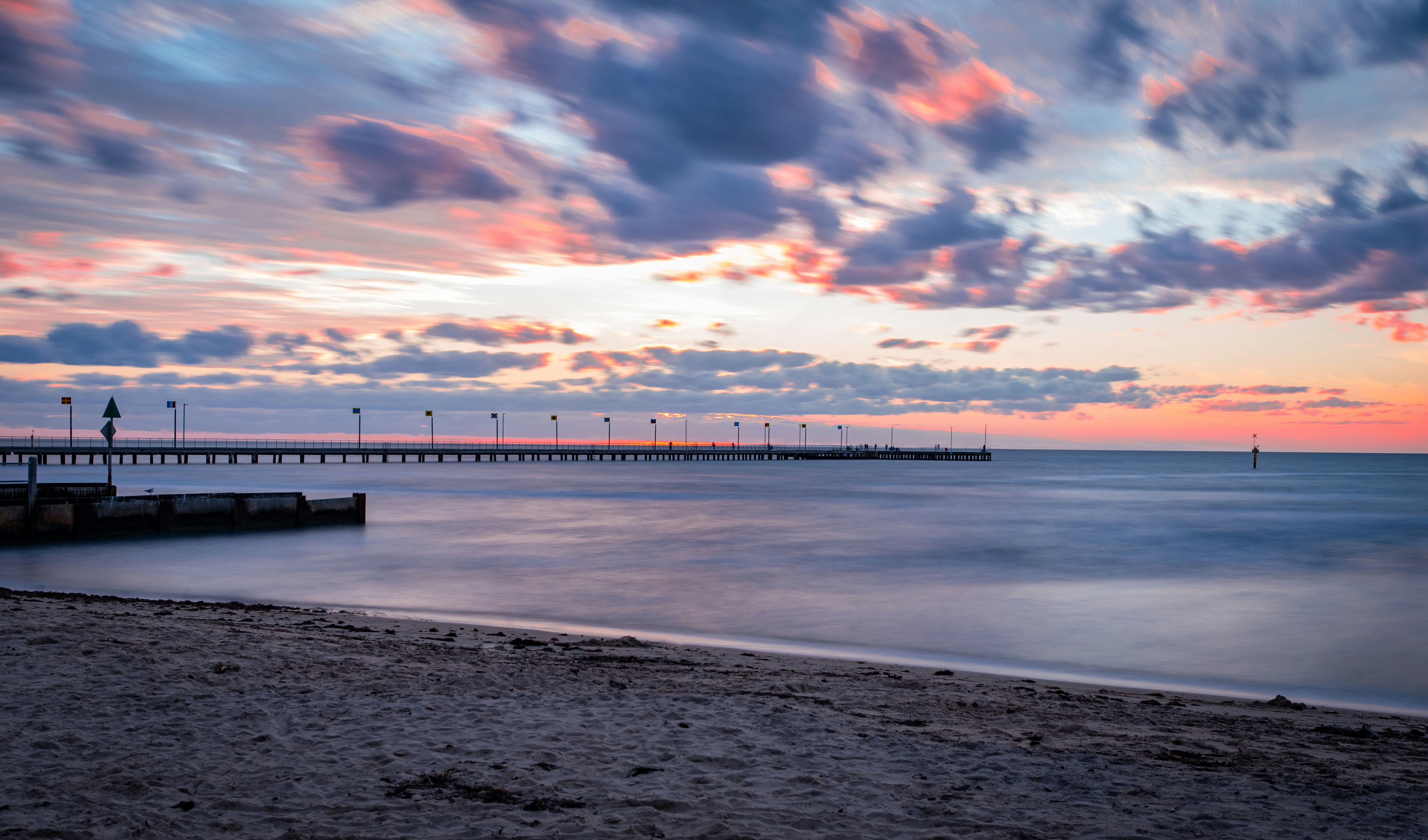 Serene beach scene at dusk, featuring a long pier extending into the calm sea under a colorful sky filled with clouds. 