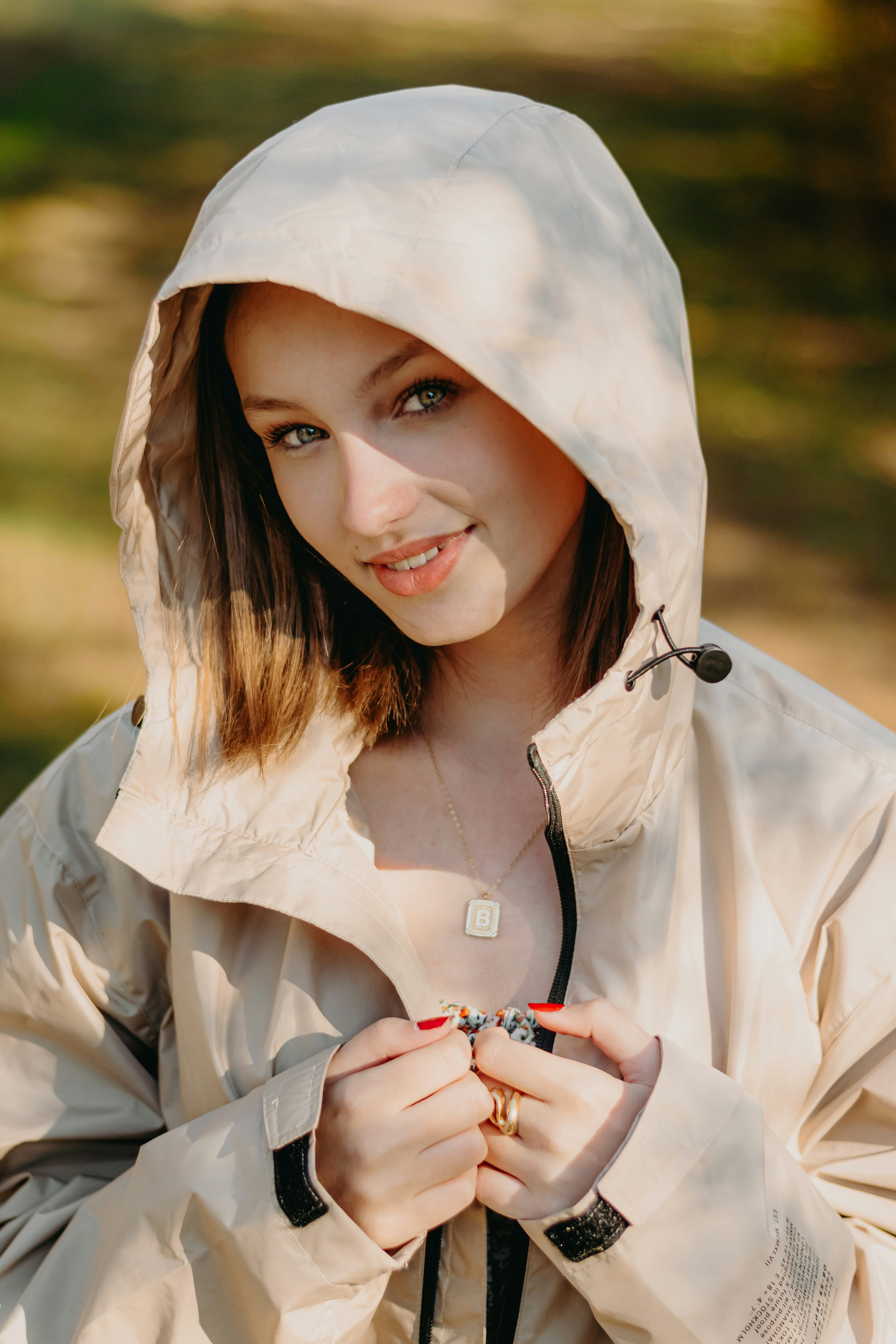 Young woman in a light jacket, smiling softly while holding her hands together, surrounded by a natural backdrop.