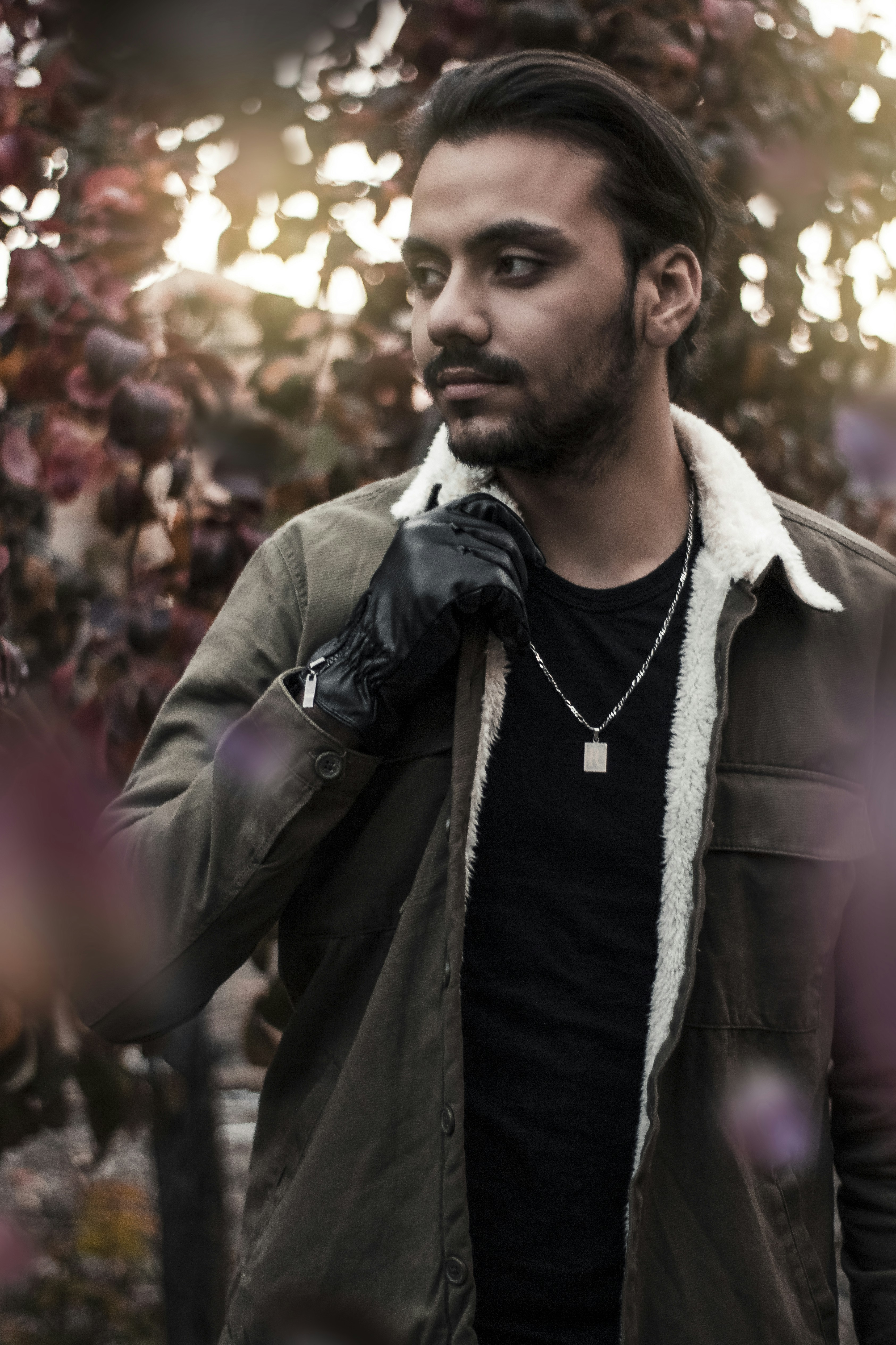 A man in a stylish jacket and gloves stands against a backdrop of autumn foliage, exuding a sense of urban sophistication.