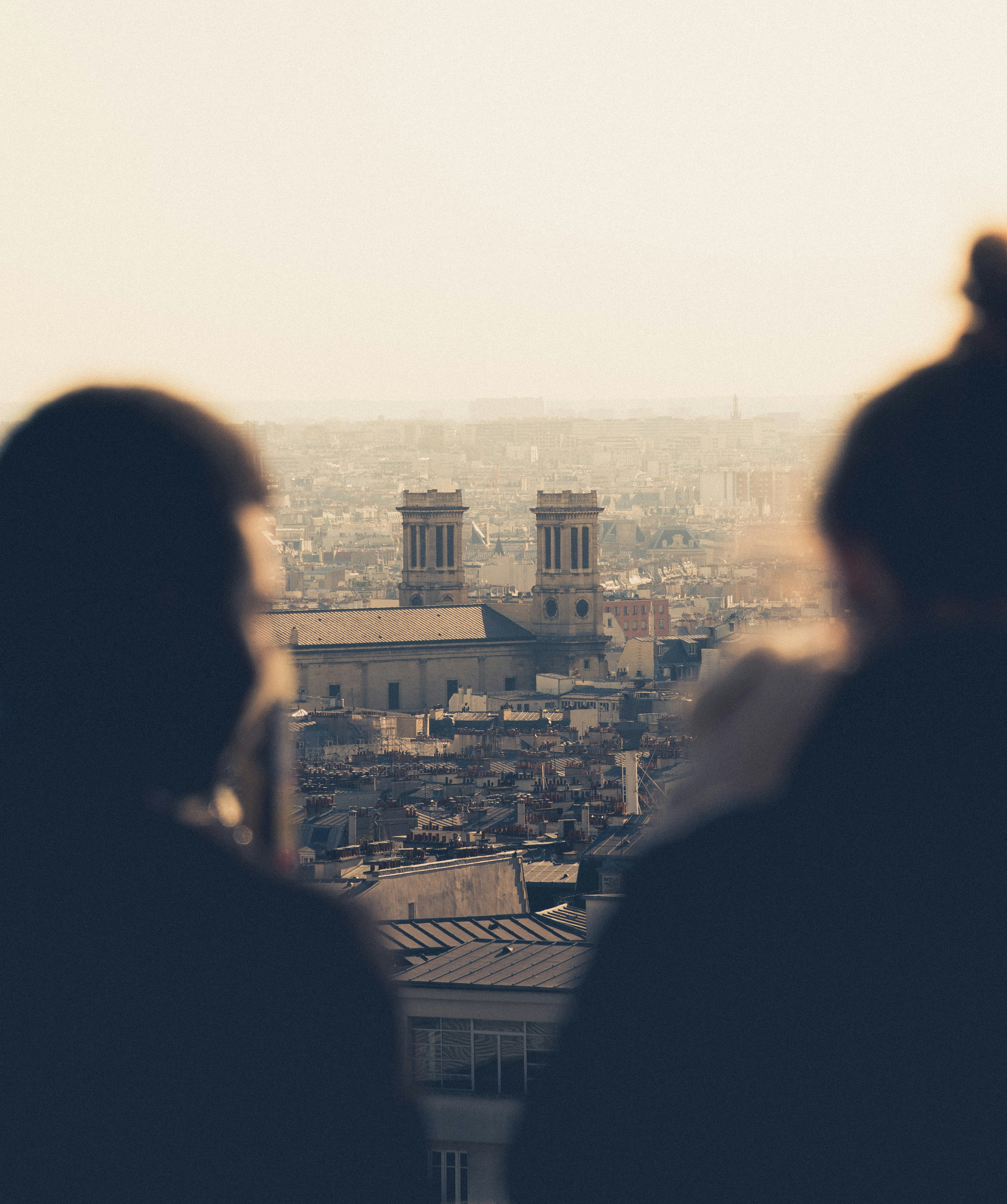 Two silhouettes overlook a sprawling urban landscape, with historic architecture peeking through the hazy atmosphere.