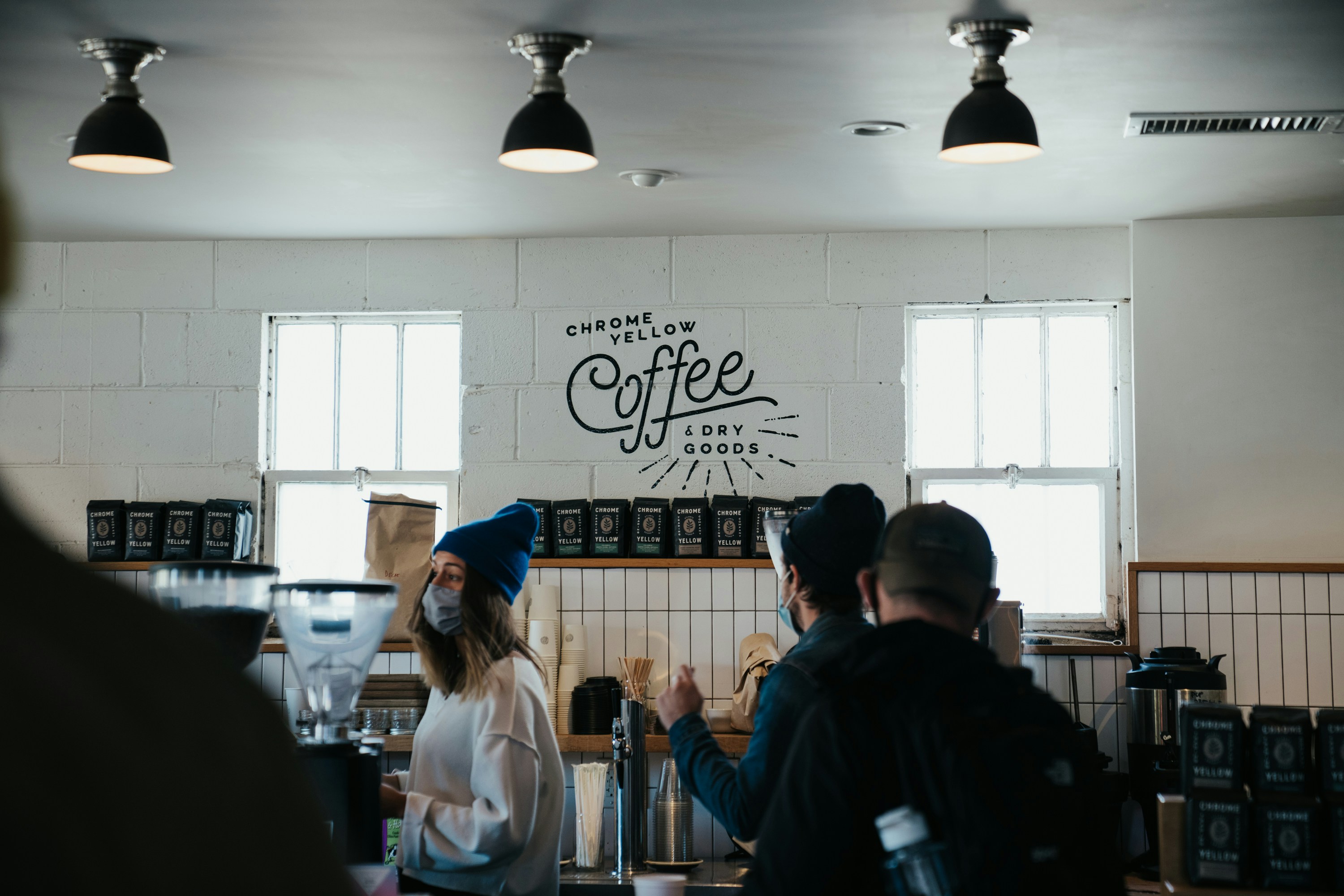 Busy coffee shop interior with people gathered at the counter beneath vintage-style lighting.