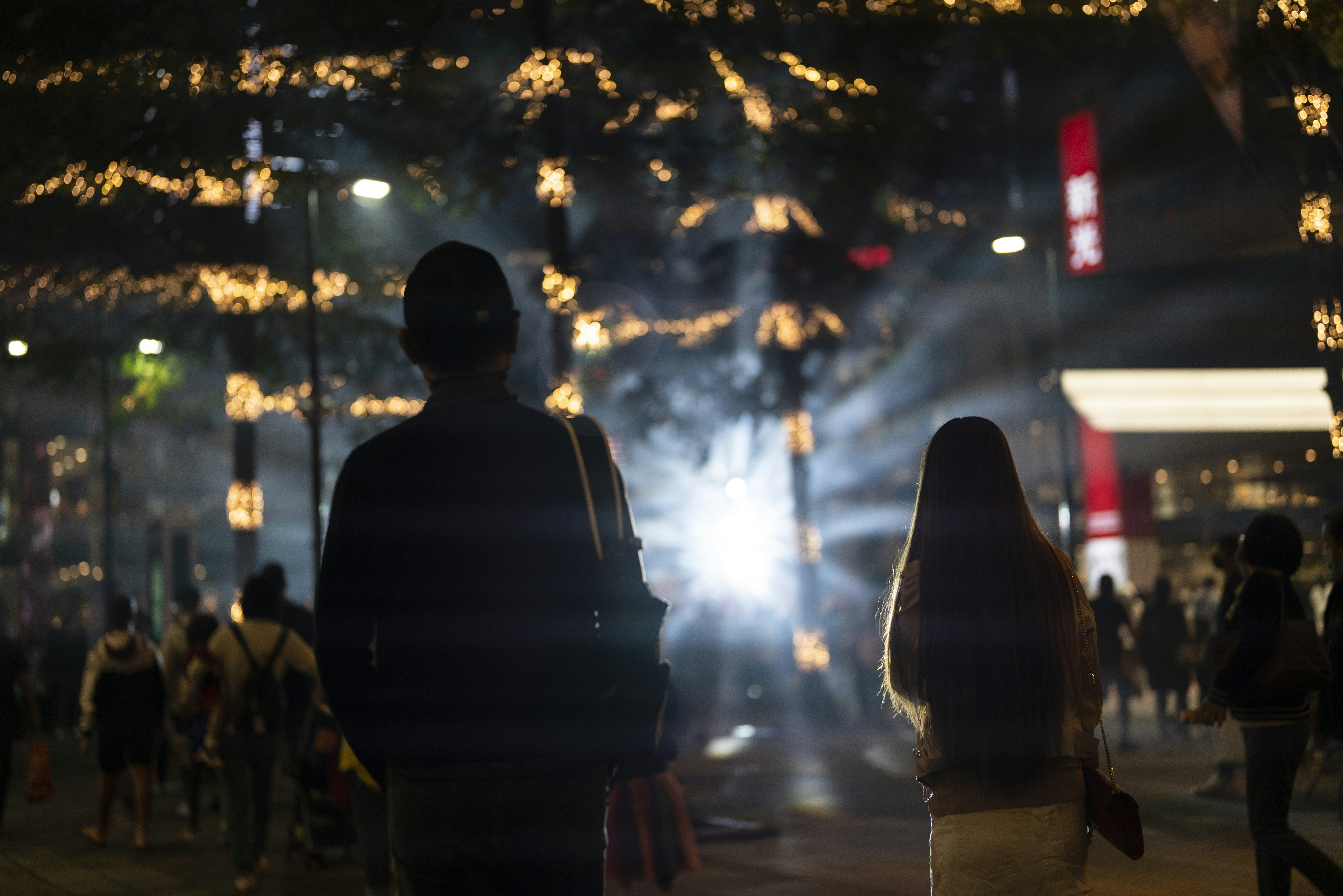 Silhouetted figures stand amidst glowing lights and mist, creating an enchanting atmosphere in an urban setting.