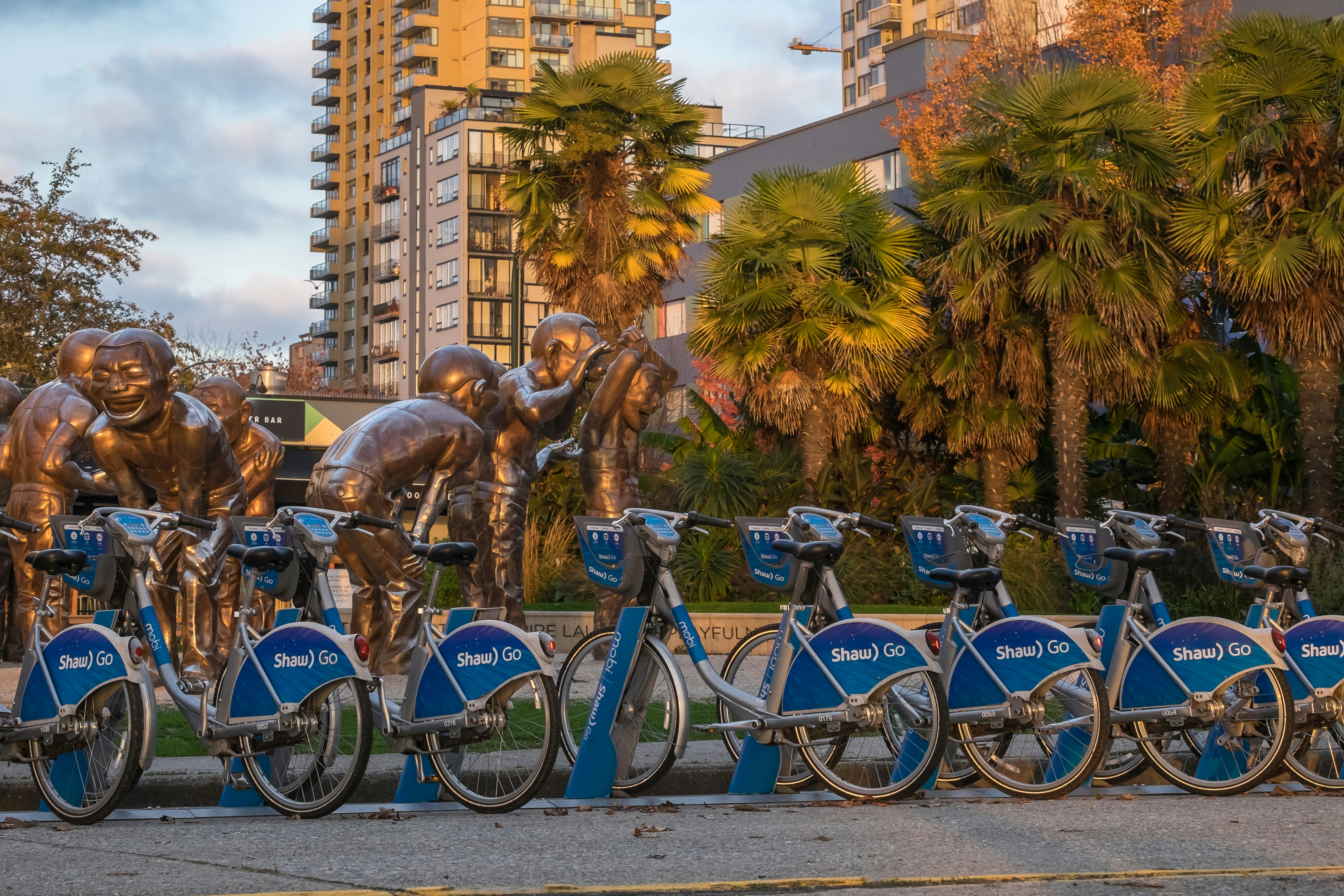 Bronze statues of playful children interact beside a row of blue rental bicycles in an urban park setting. 