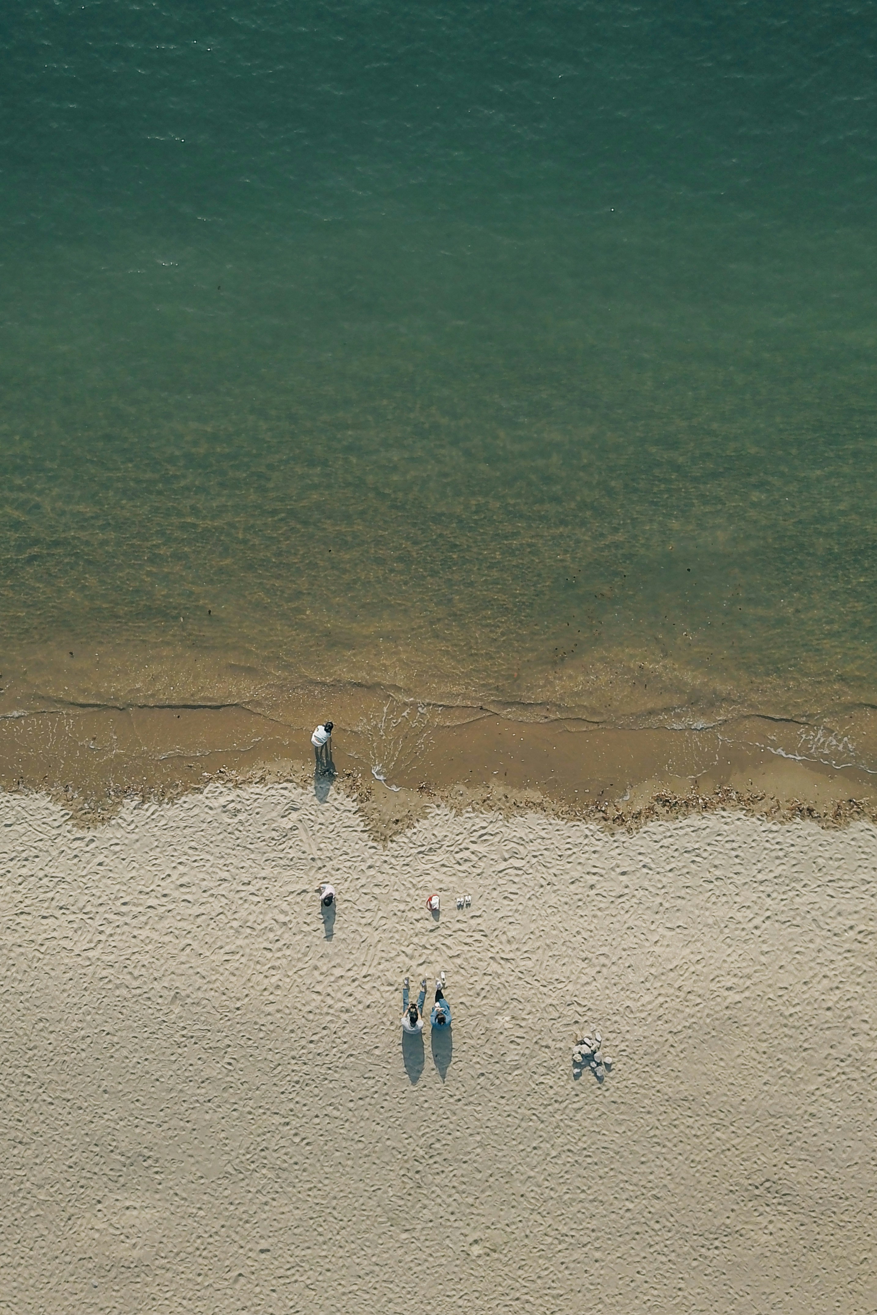 Aerial view of people enjoying a sunny day on the beach, with gentle waves lapping at the shore. The scene captures the essence of leisure and tranquility.