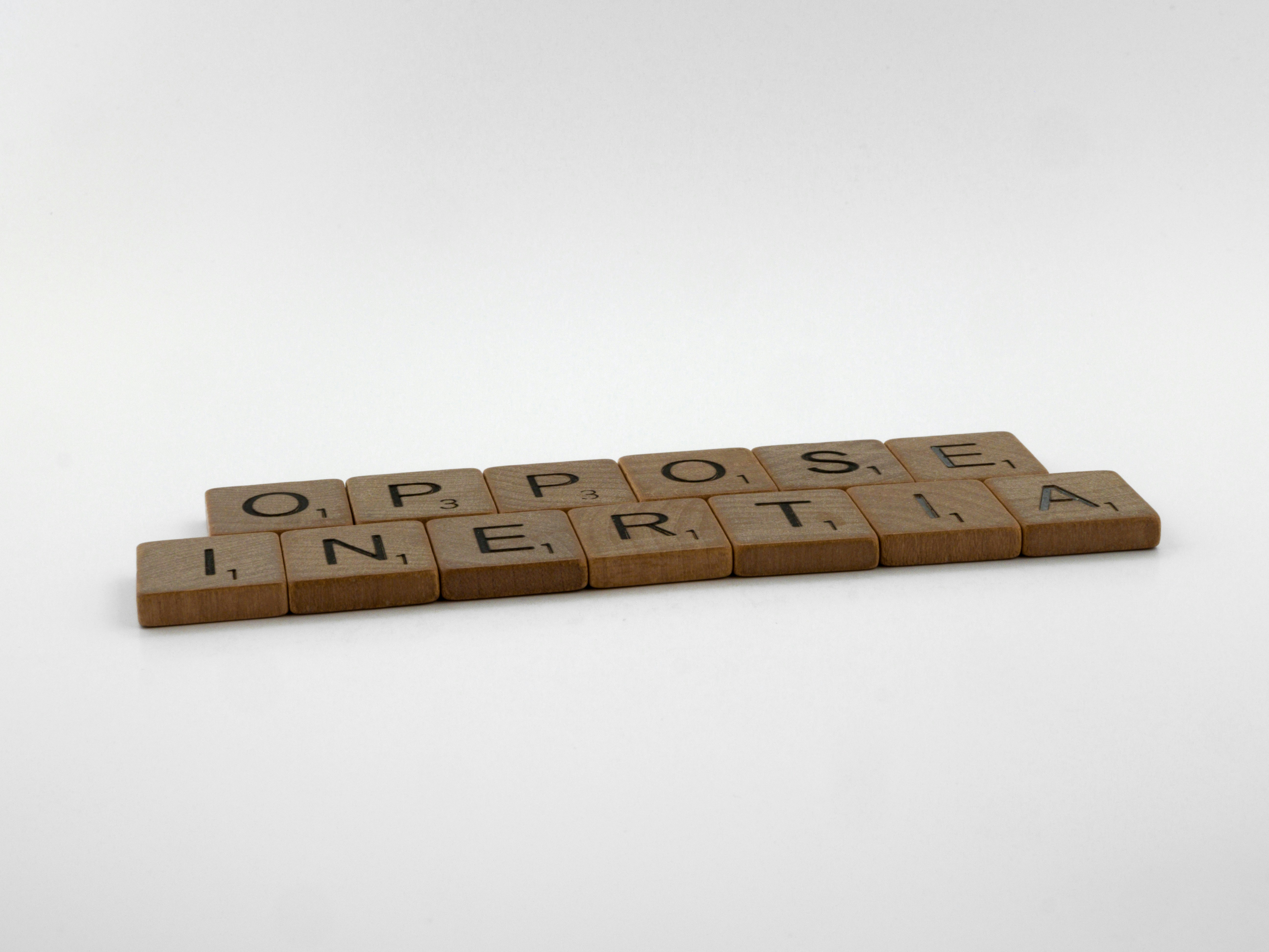 Wooden letter tiles arranged to spell 'OPPOSE INERTIA' against a neutral background.