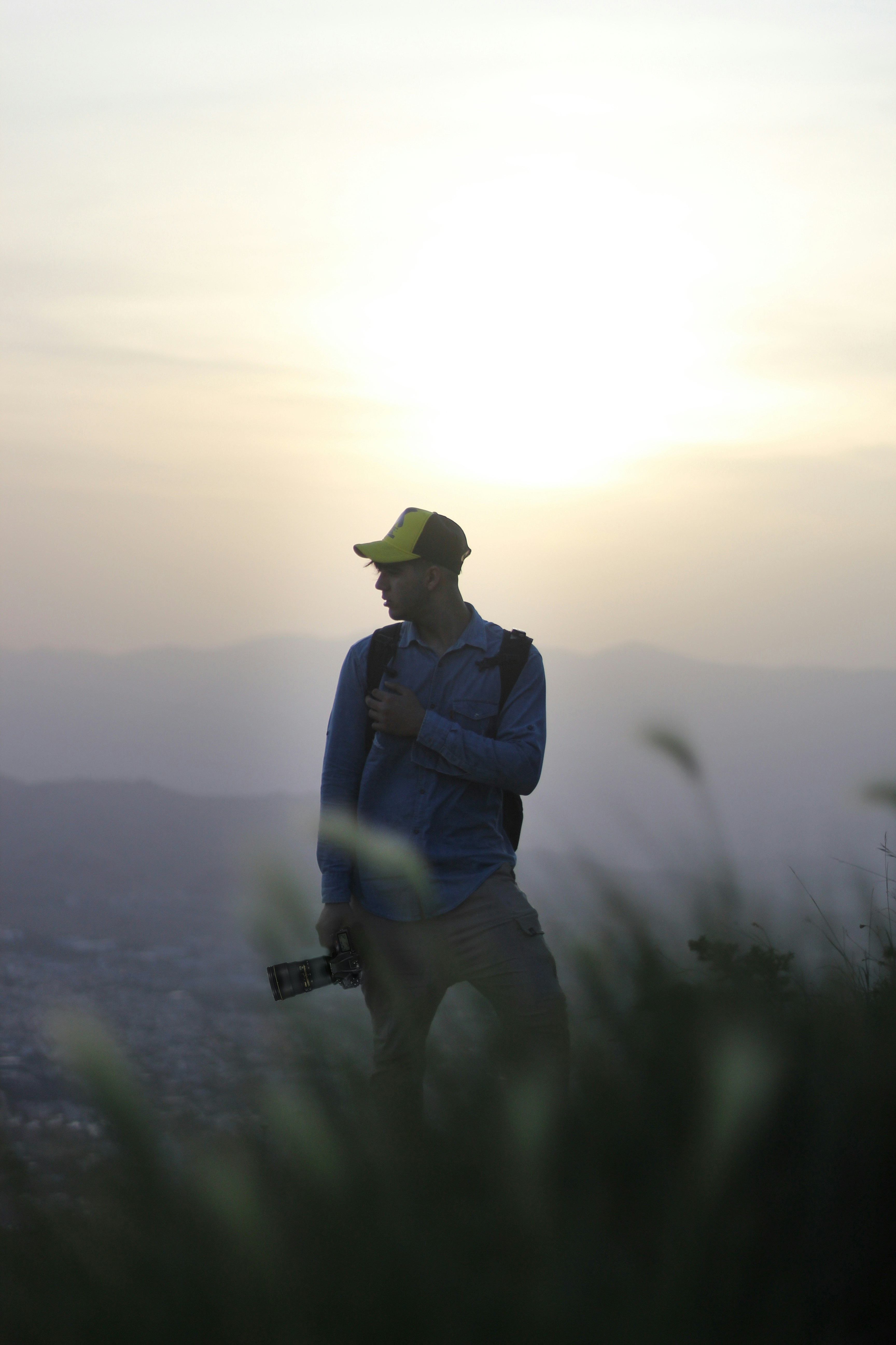 Silhouette of a photographer standing on a hilltop, holding a camera against a soft, glowing sunset. The surrounding landscape fades into the horizon.