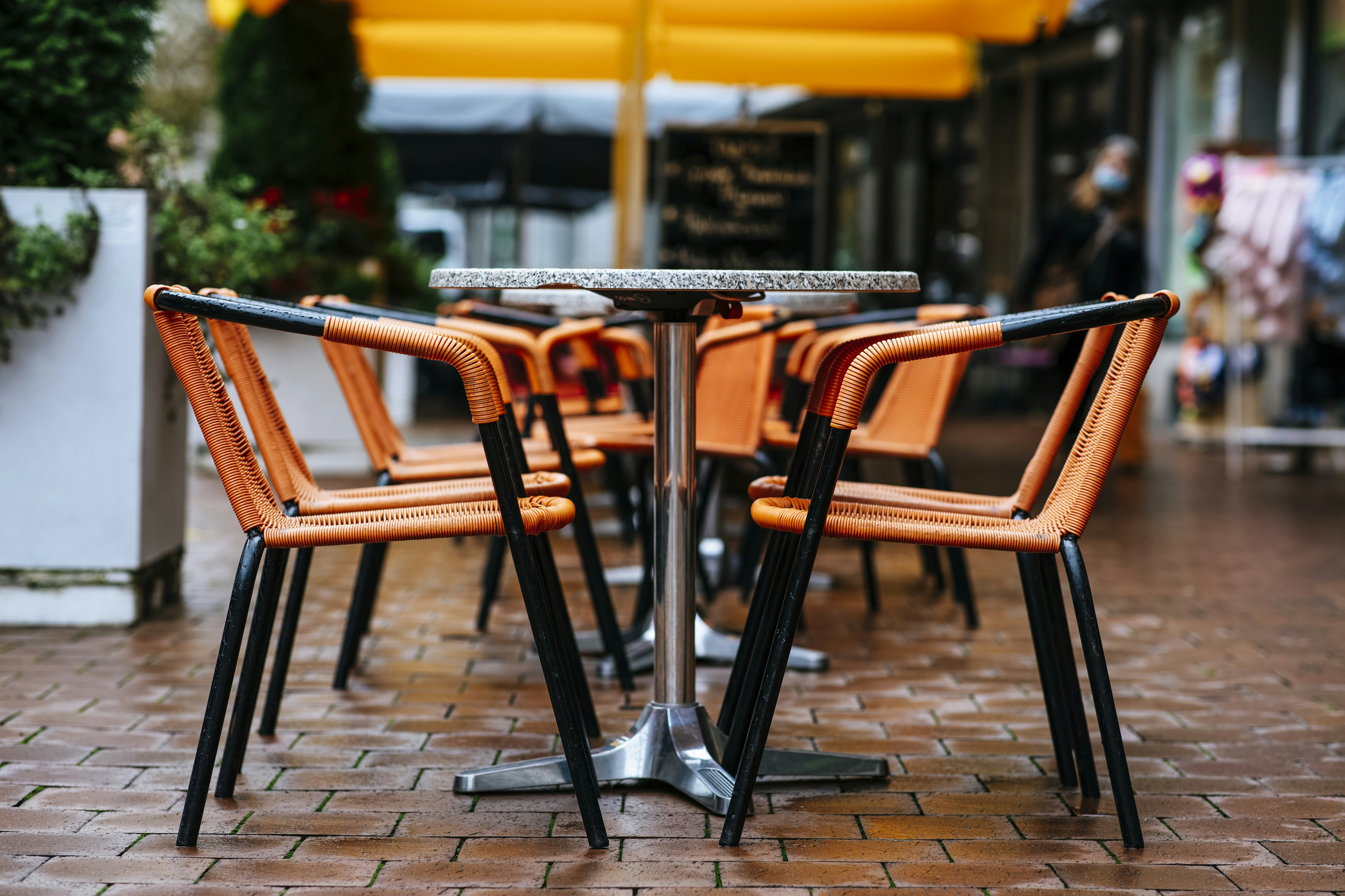 Empty orange and black chairs arranged around a small table under a vibrant yellow umbrella in a bustling outdoor cafe setting.